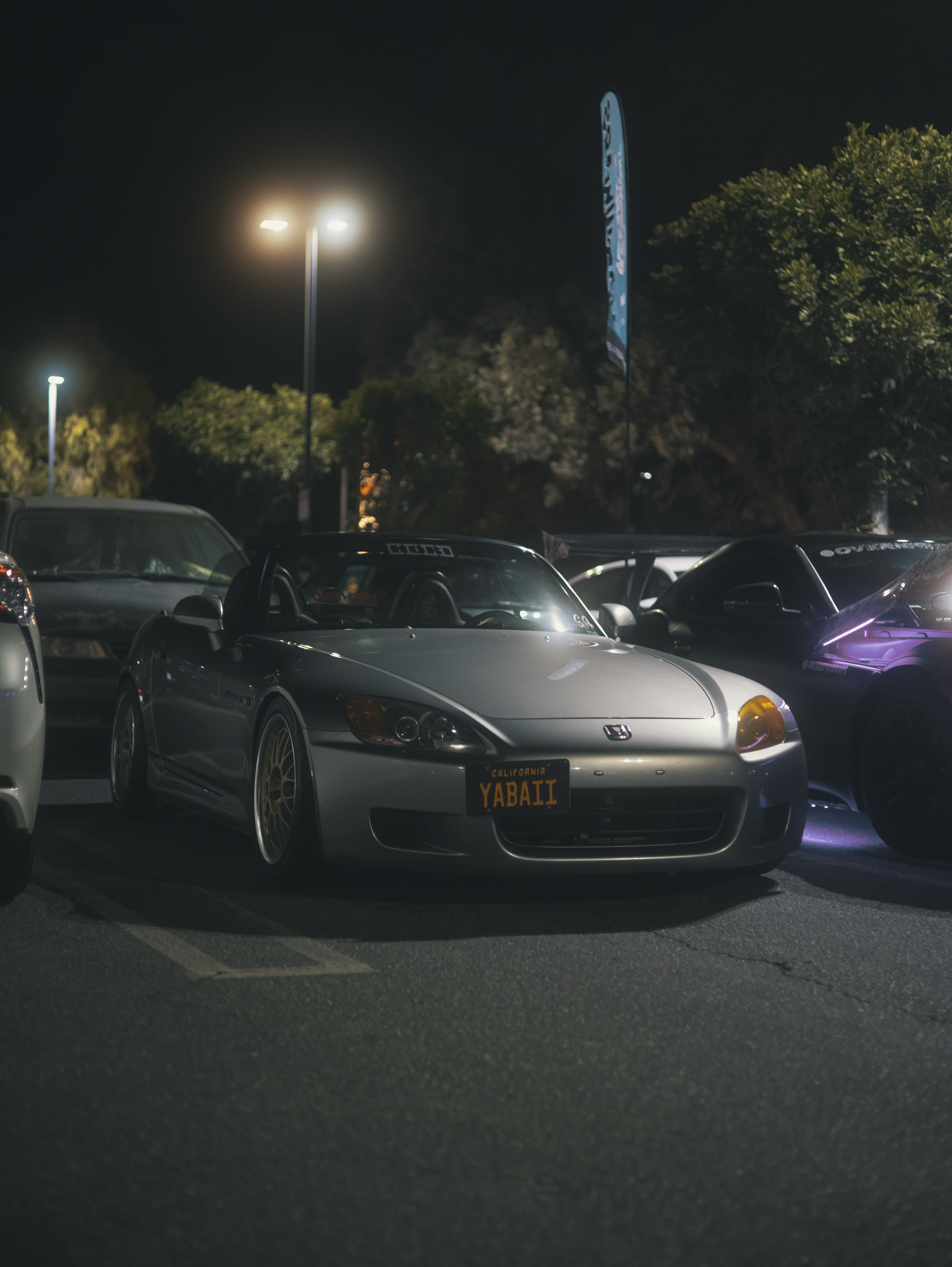 Silver Honda S2000 parked at night among other cars, illuminated by streetlights. The scene captures the essence of car culture and nighttime gatherings.