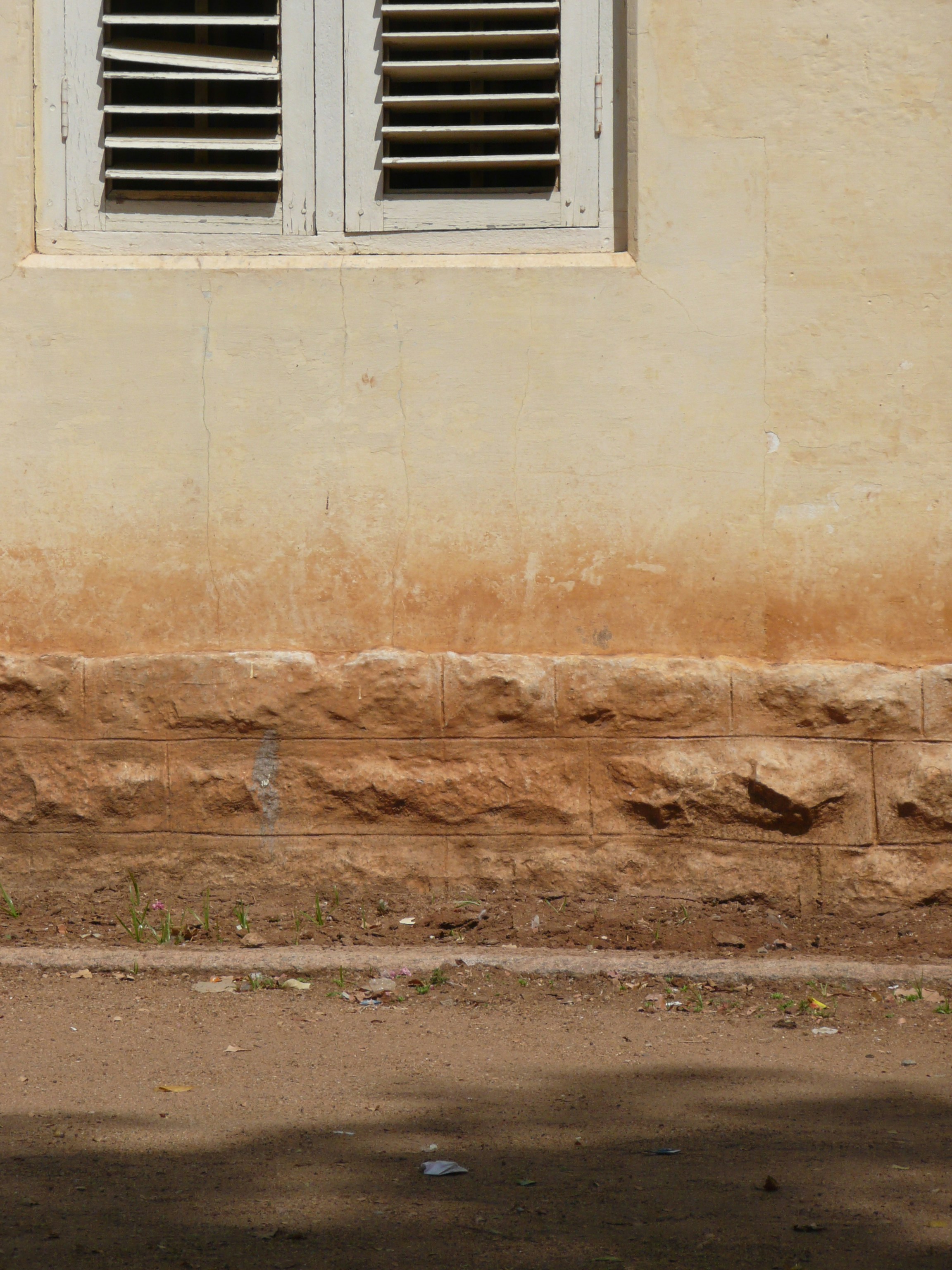 Two weathered louvered shutters sit on a sunlit beige wall above a rough stone base, with a shadowed ground in the foreground.