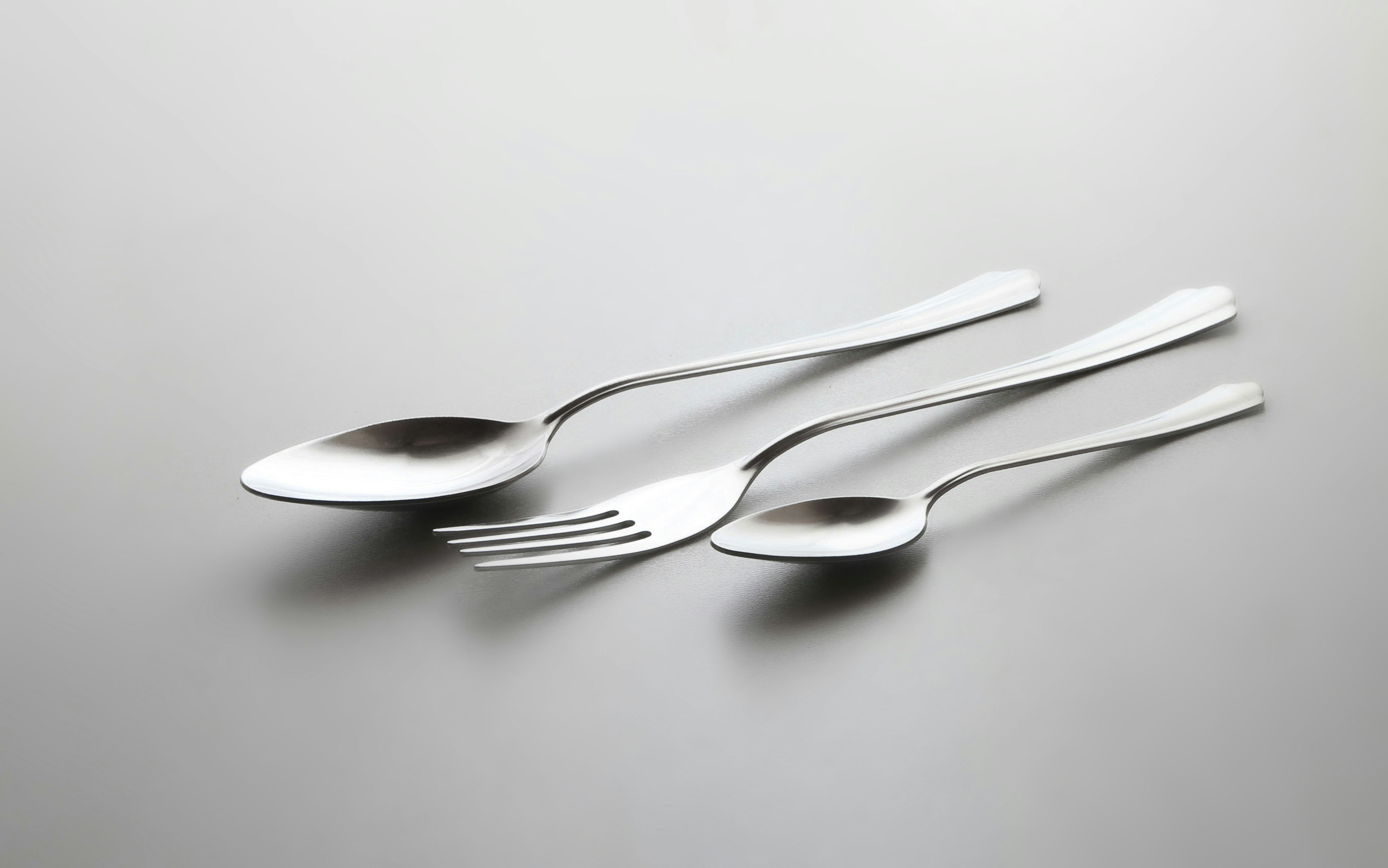 Three elegantly arranged stainless steel utensils—spoon, fork, and teaspoon—set against a soft, neutral background.