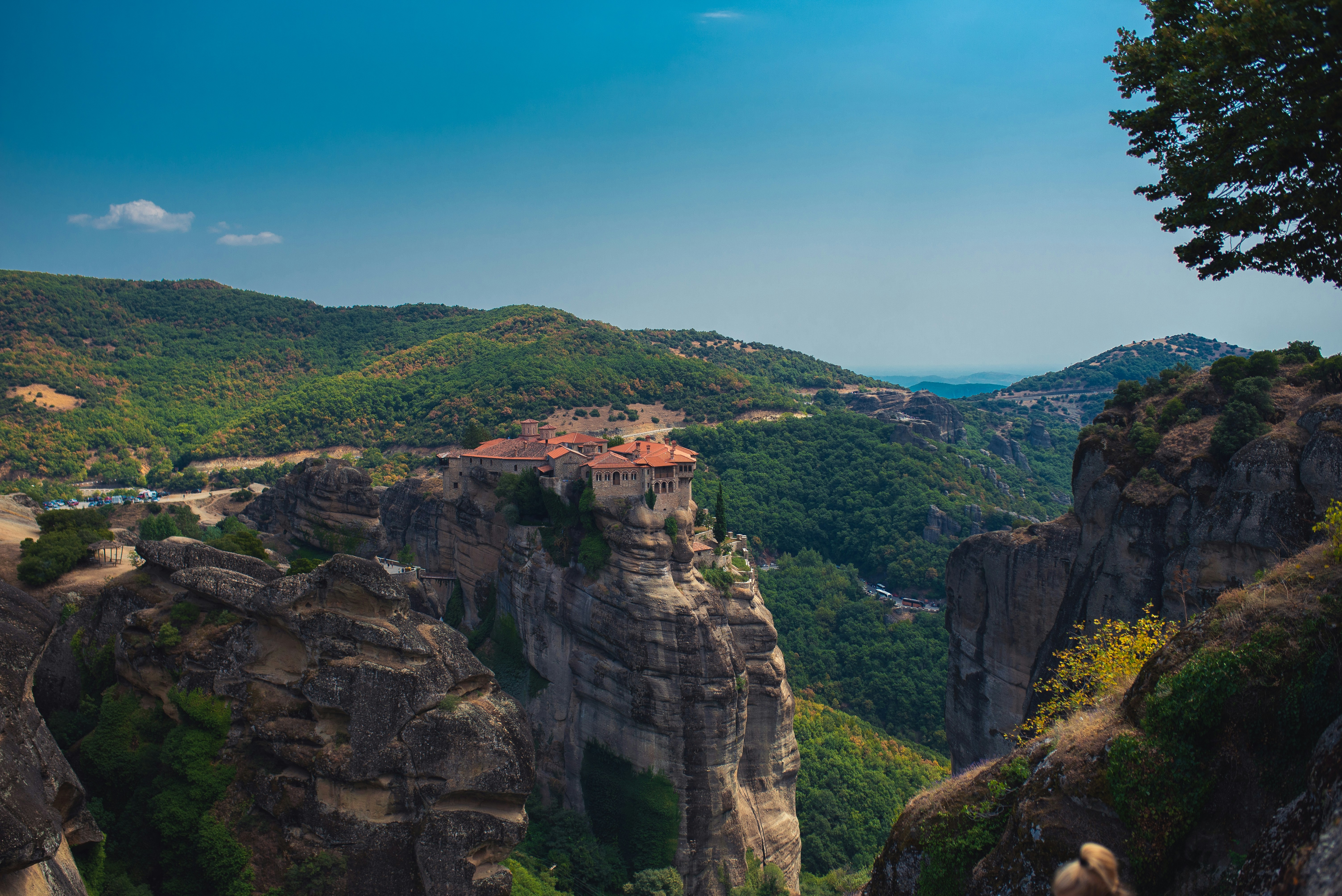 Ancient monastery atop towering rock formations surrounded by lush green hills under a clear blue sky.