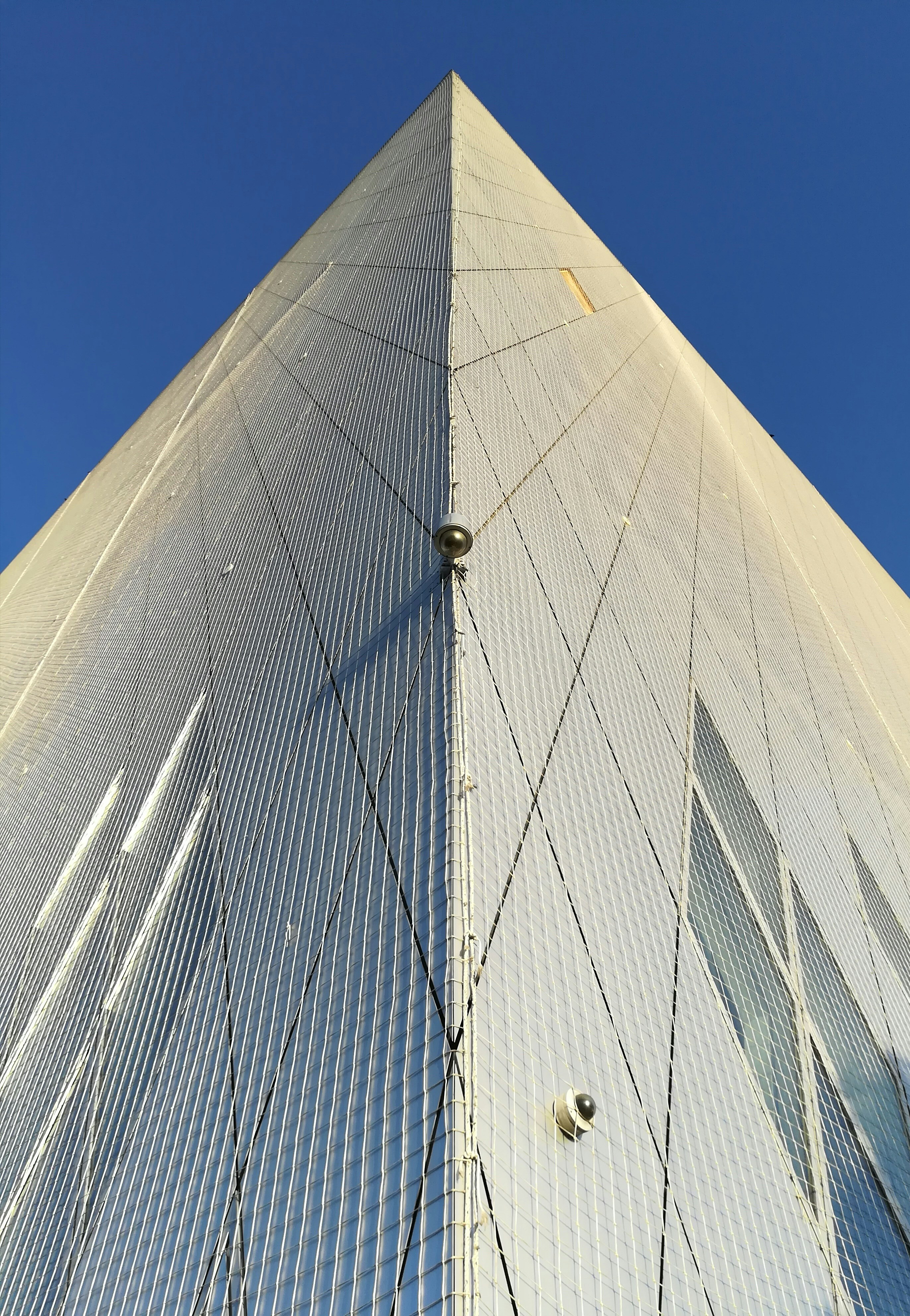A tall glass-clad spire with a diamond-pattern facade rises toward a clear blue sky. This photograph emphasizes converging lines and the reflective surface of the building.