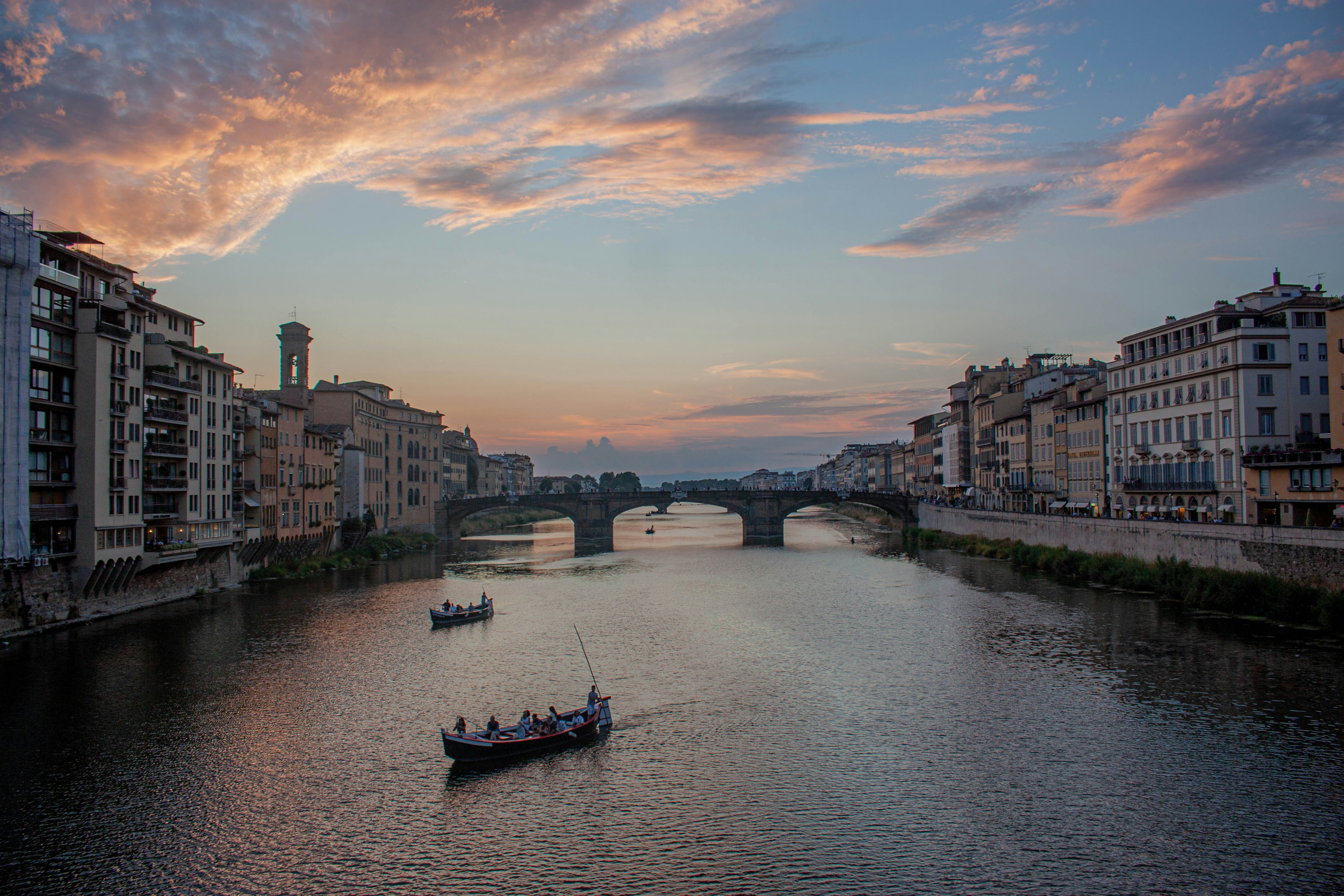 Serene view of the Arno River with boats drifting under historic bridges at dusk, framed by charming architecture and a colorful sky.