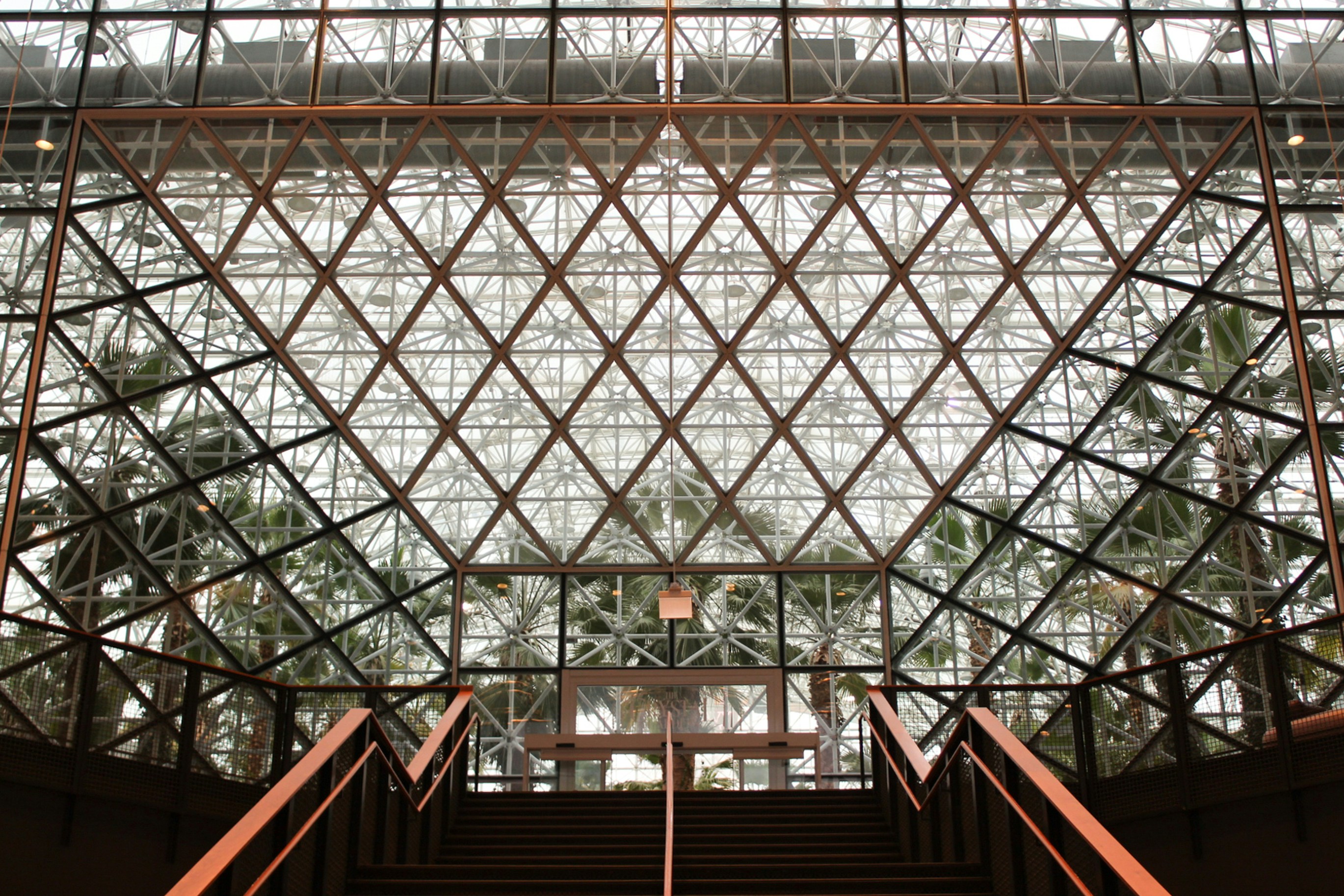 Intricate glass and metal structure showcasing a modern architectural design with a view of palm trees through the transparent ceiling.