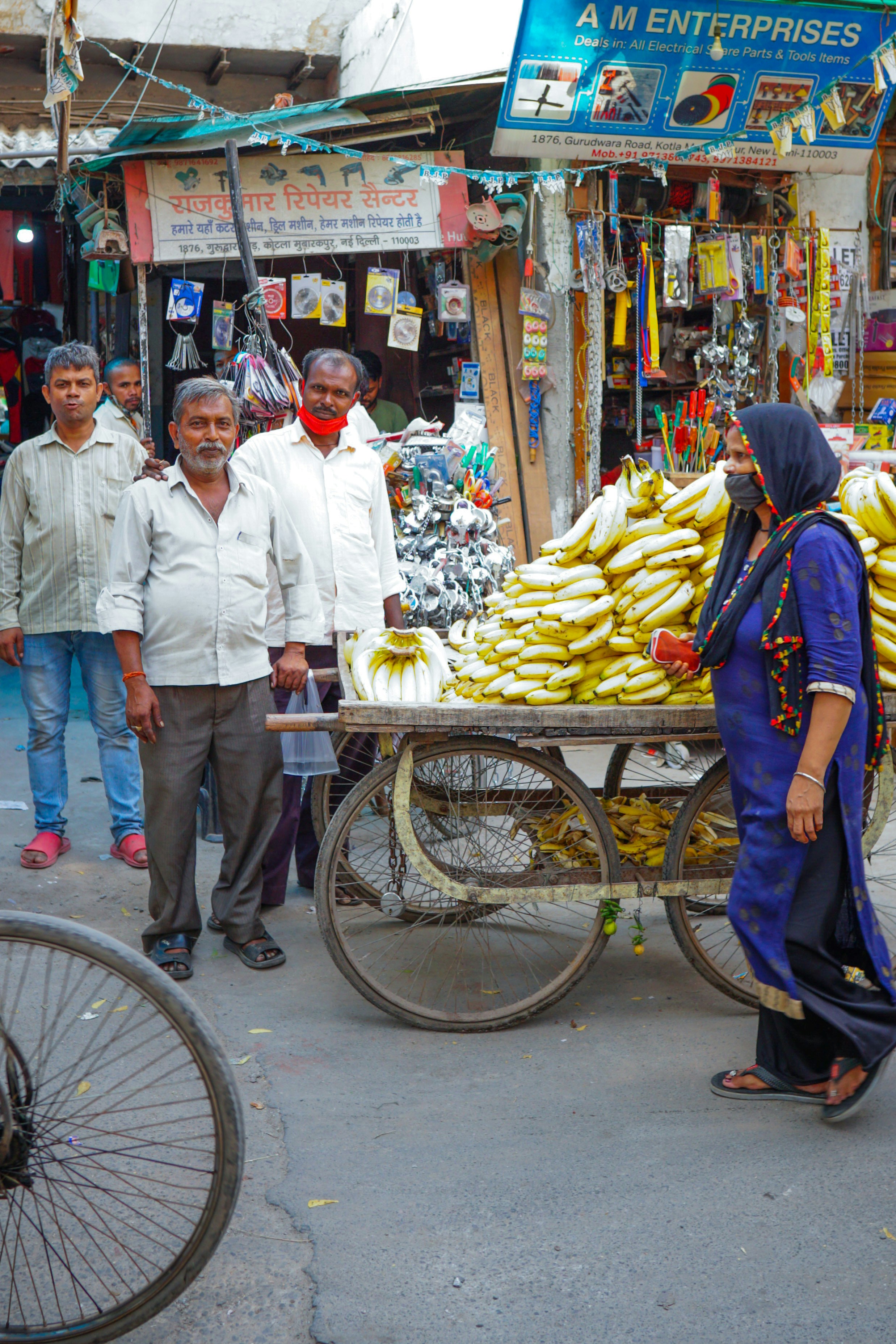 A street vendor with a cart full of bananas interacts with a passerby in a busy Delhi market.