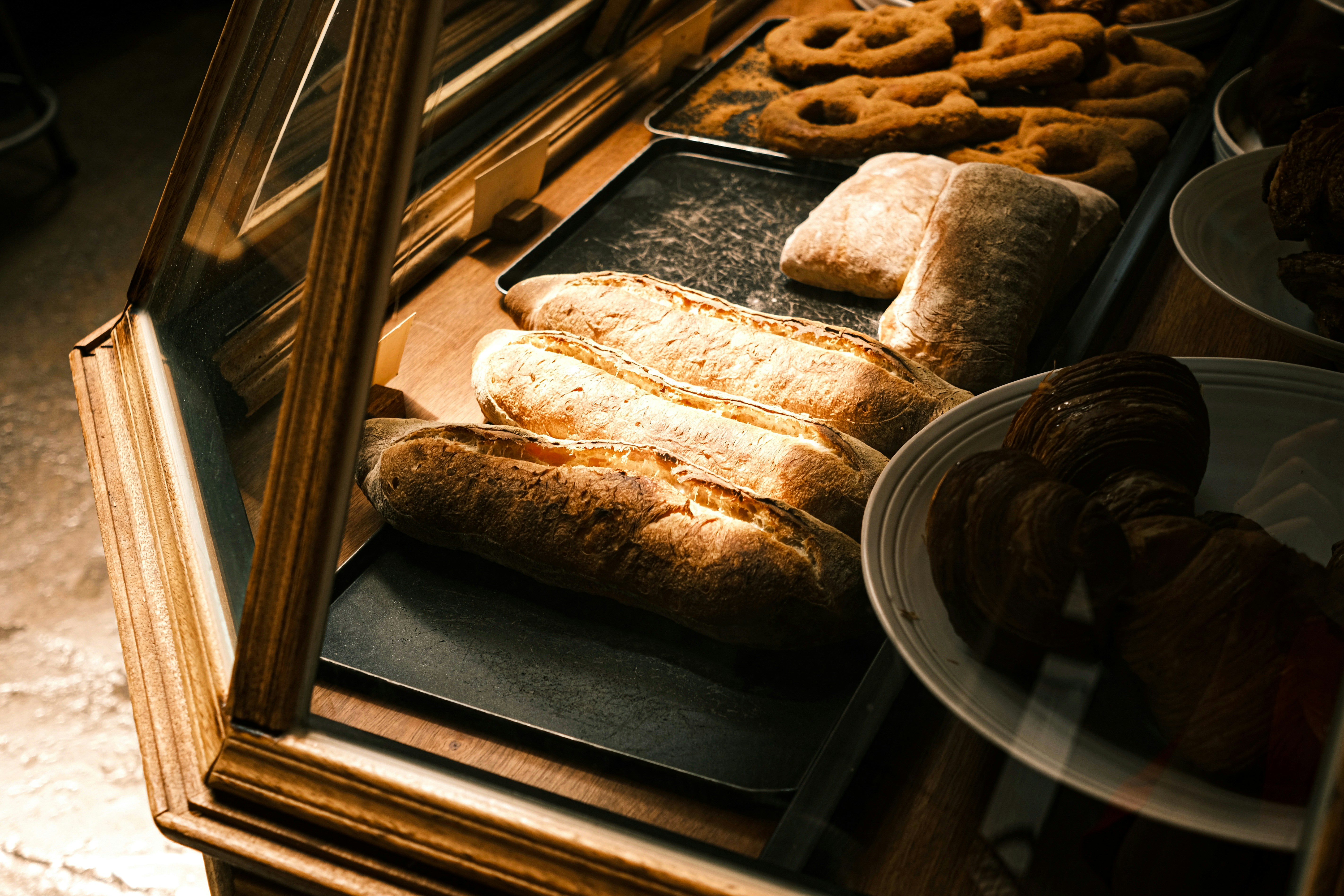 Freshly baked artisan breads displayed under a glass case, highlighting their textures and colors. The warm lighting enhances the inviting atmosphere.