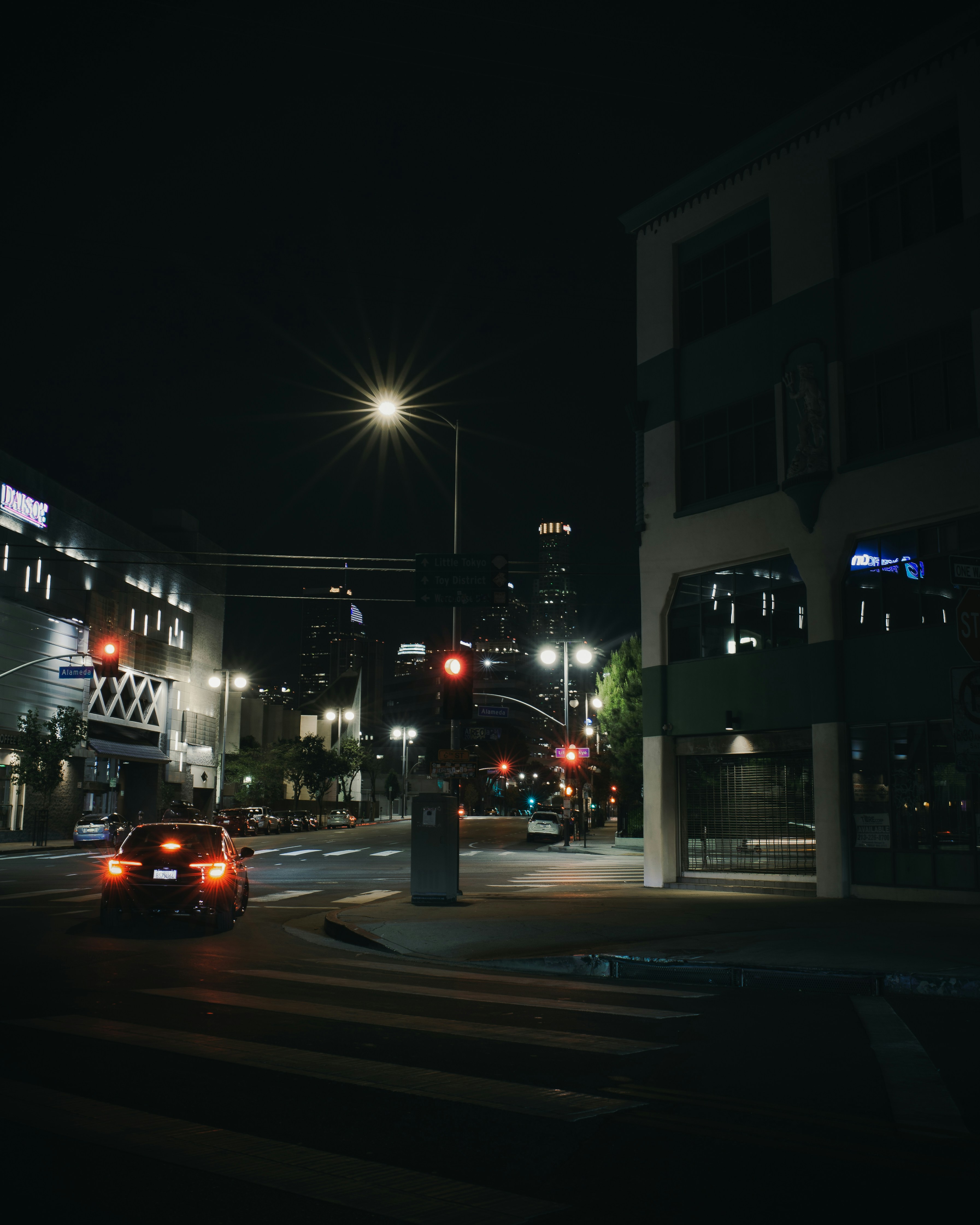 A car navigates a quiet city intersection illuminated by streetlights, with distant skyscrapers glowing under the night sky.