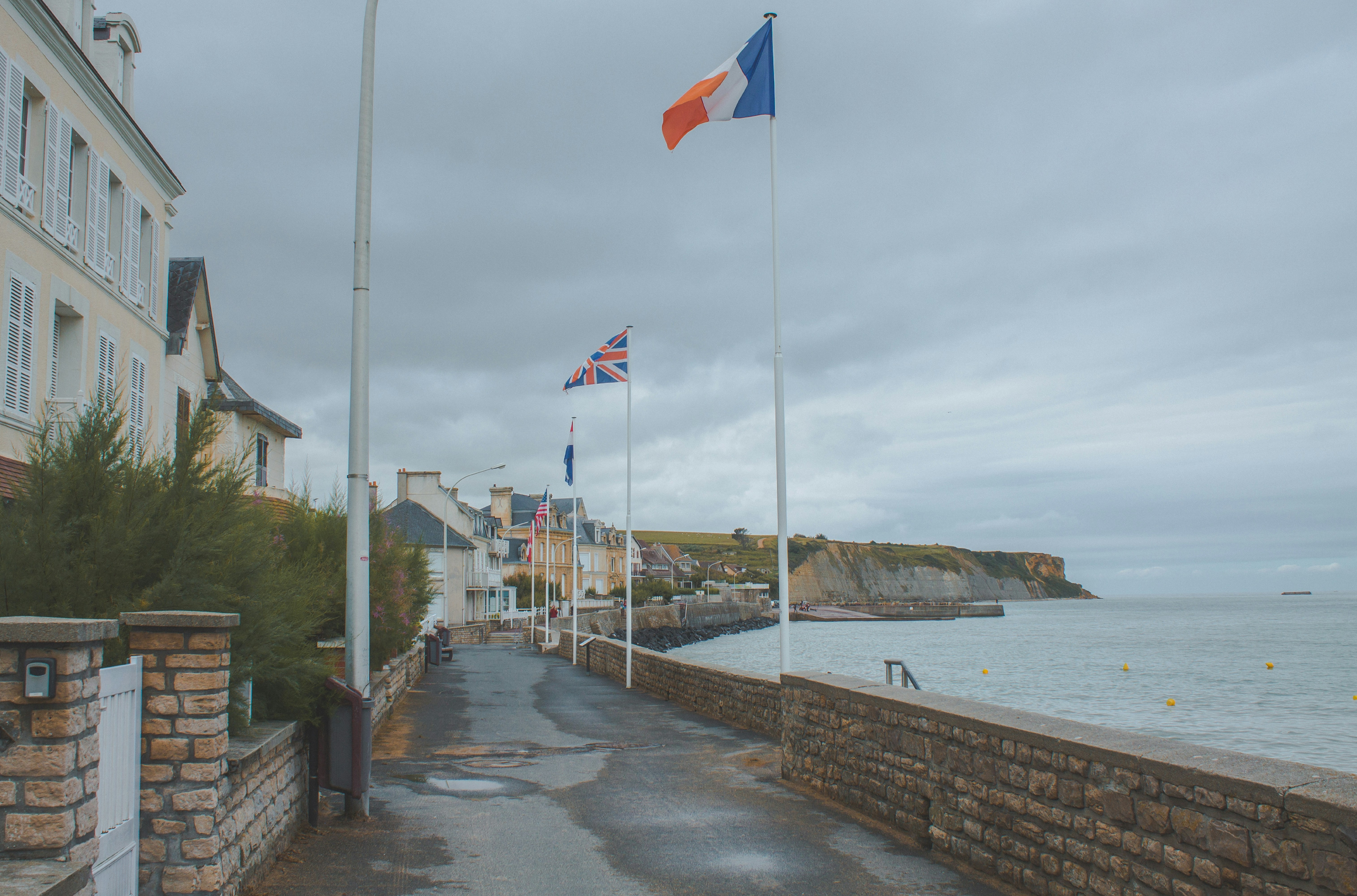 Coastal walkway lined with flags representing France and the UK, leading towards rocky cliffs under an overcast sky.
