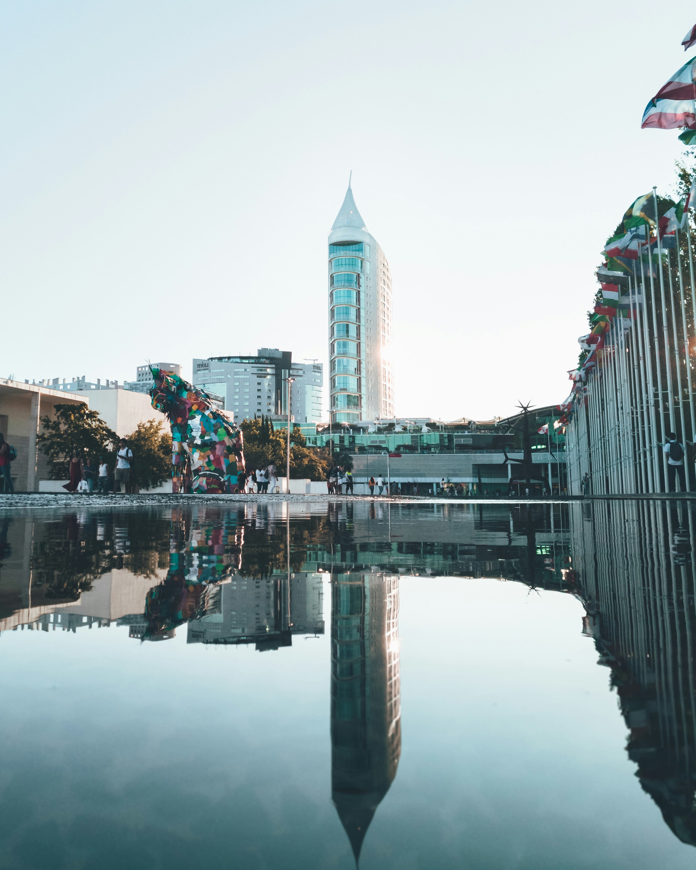 Tall building reflected in a calm, mirror-like water surface, flanked by colorful urban structures and flags at sunrise.