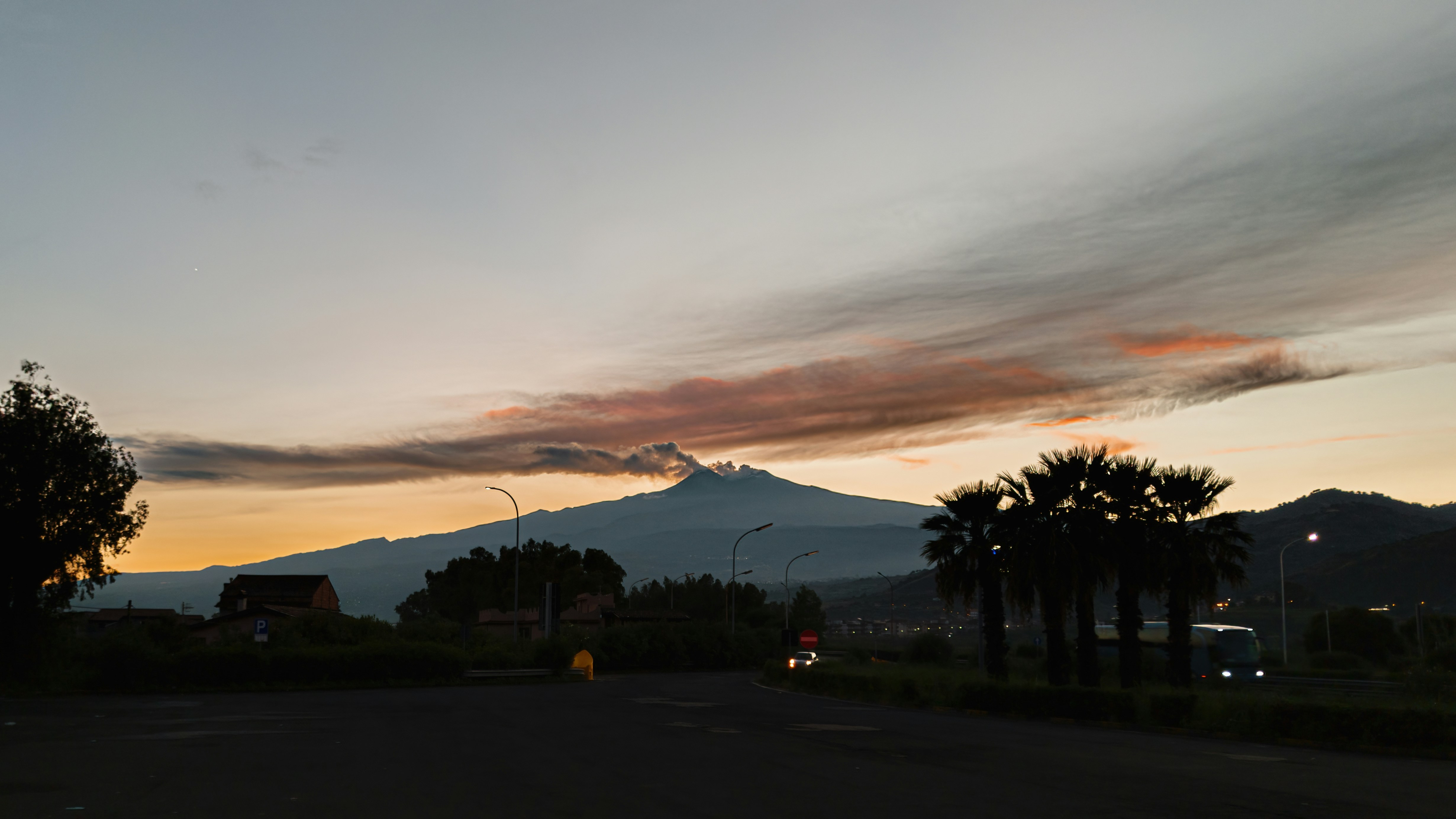 Dusk settles over a distant volcano ridge, silhouetted against a gradient sky. Palm trees frame the right side as a quiet road anchors the foreground.