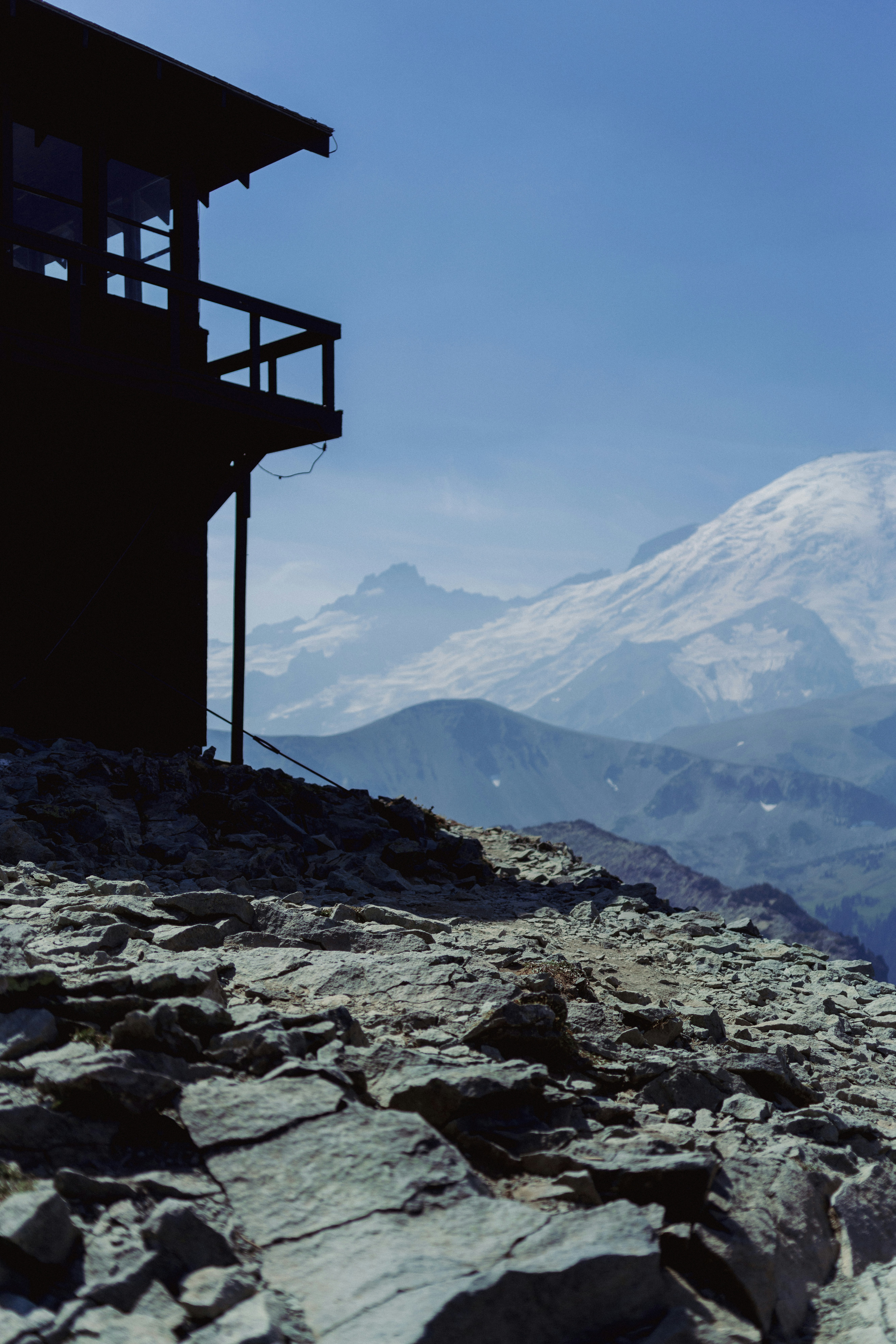 A rugged mountain landscape with a silhouette of a watchtower overlooking distant peaks and valleys. The rocky terrain leads the eye toward the majestic backdrop.