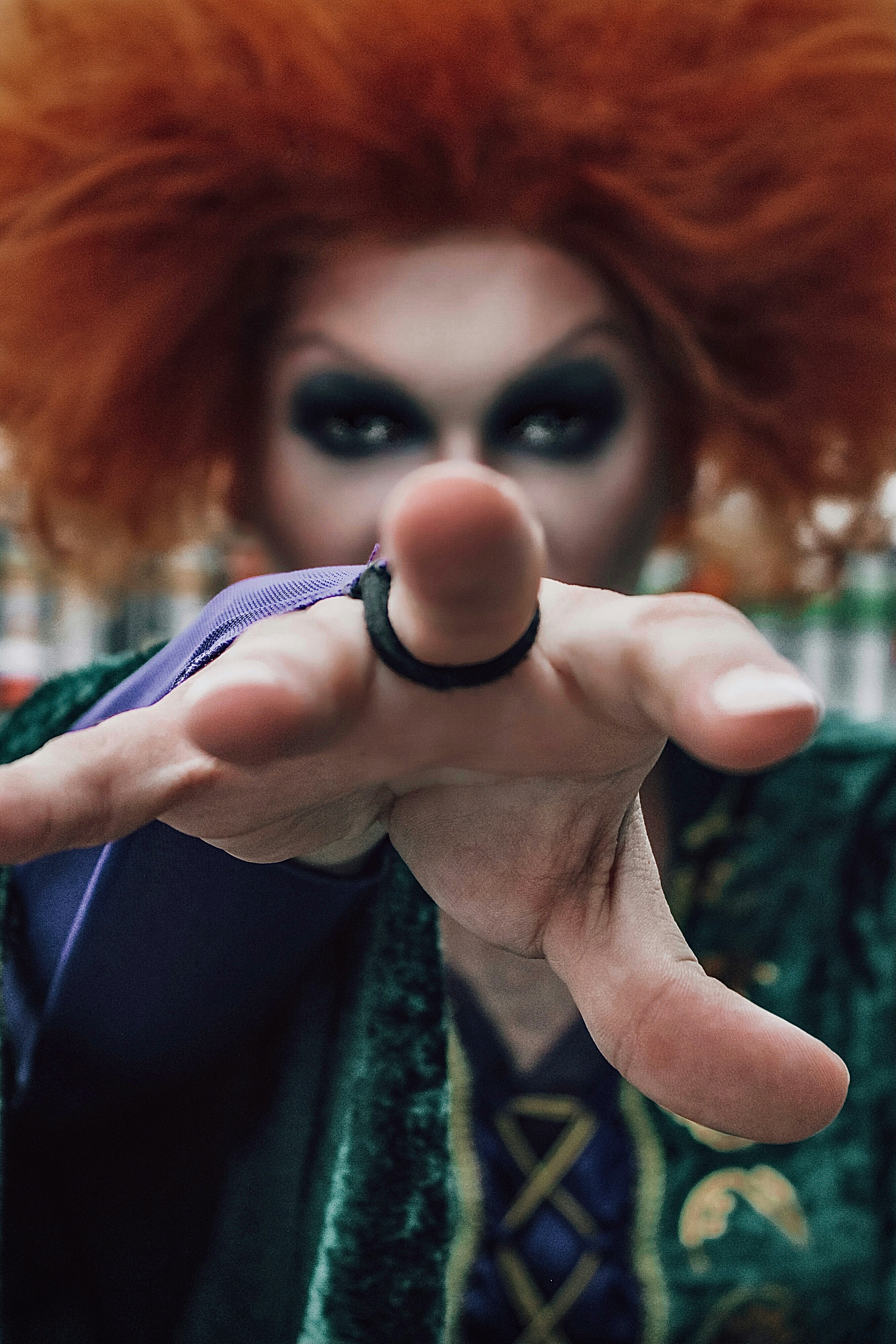 A theatrical figure extends a hand forward, adorned in vibrant costume with dramatic makeup and wild hair, evoking an aura of magic and mystery.