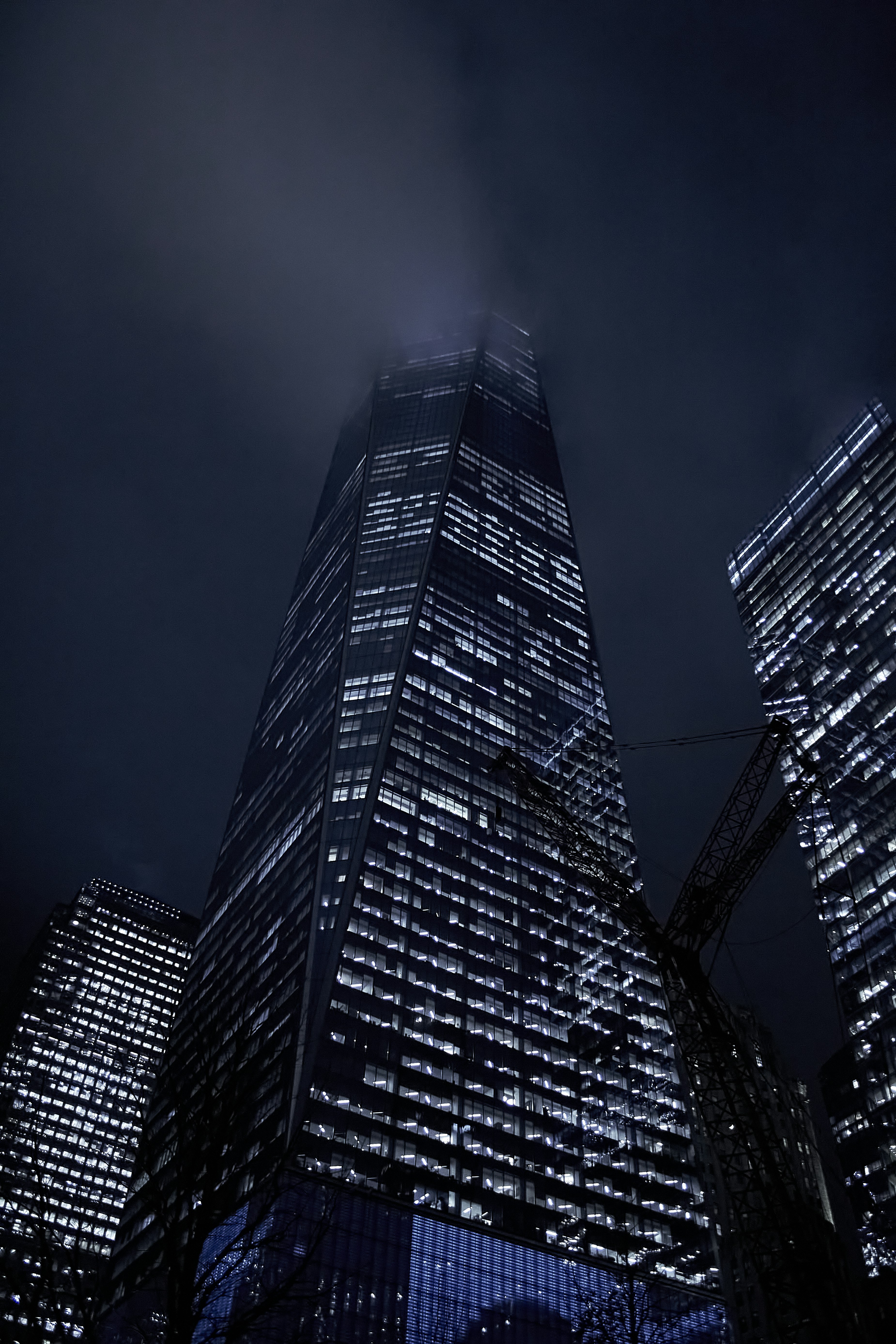 A towering skyscraper shrouded in mist, illuminated by city lights, surrounded by other buildings in a nighttime urban landscape.