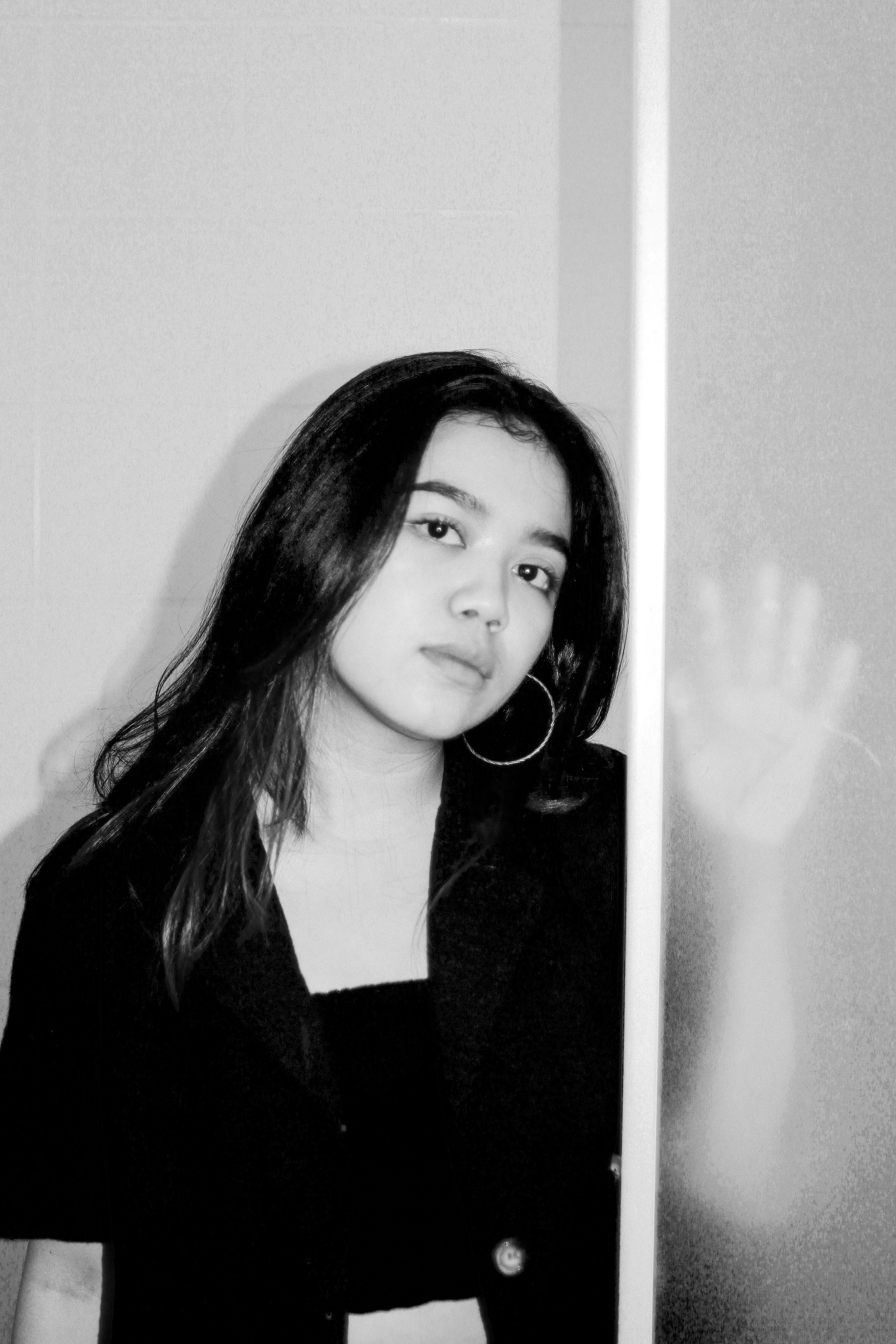 A young woman gazes thoughtfully through a translucent surface, her hand pressed against it, conveying a sense of longing. The monochrome palette enhances the emotional depth.