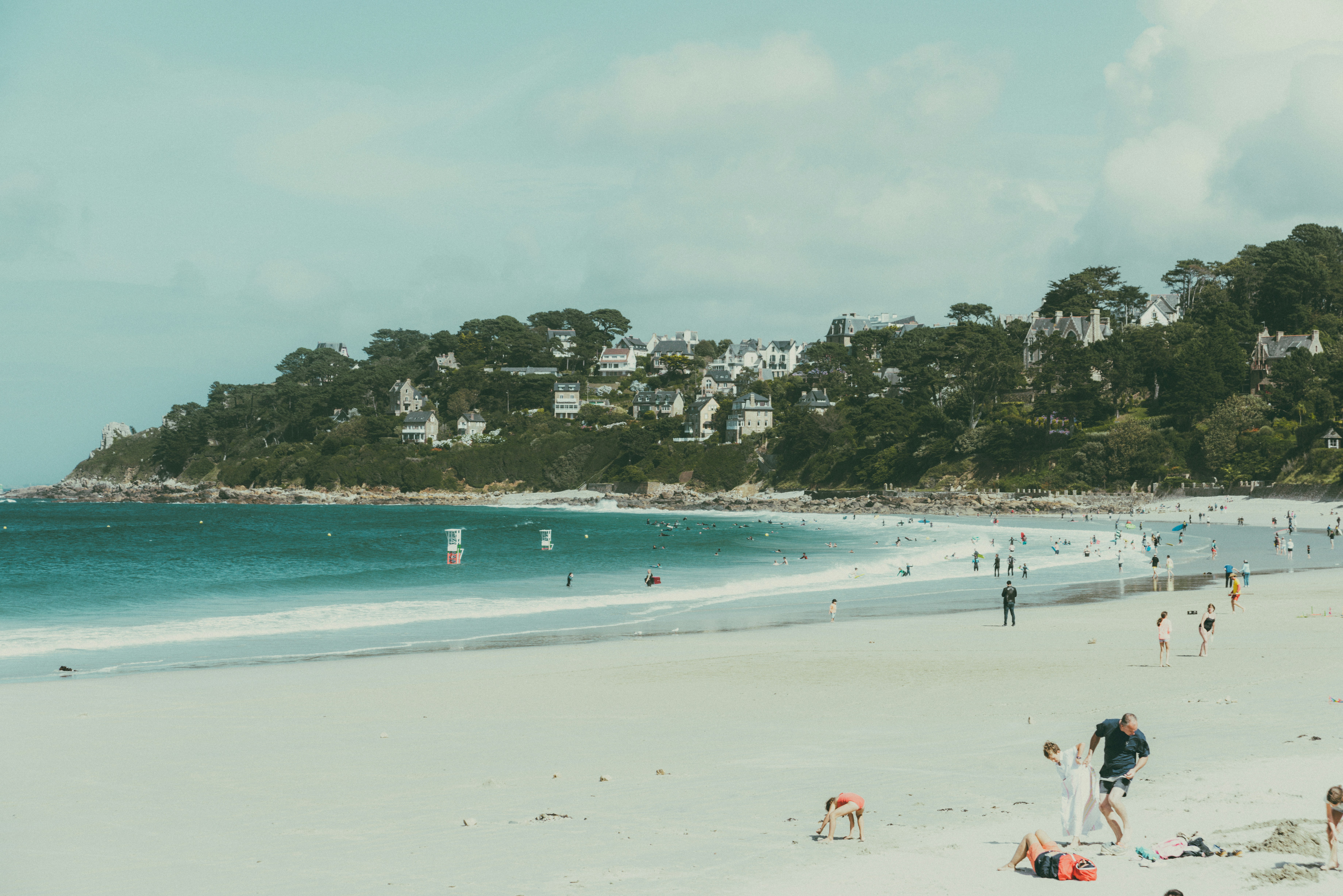 Vibrant beach scene with people enjoying the sun and water, framed by lush greenery and coastal homes in the background.