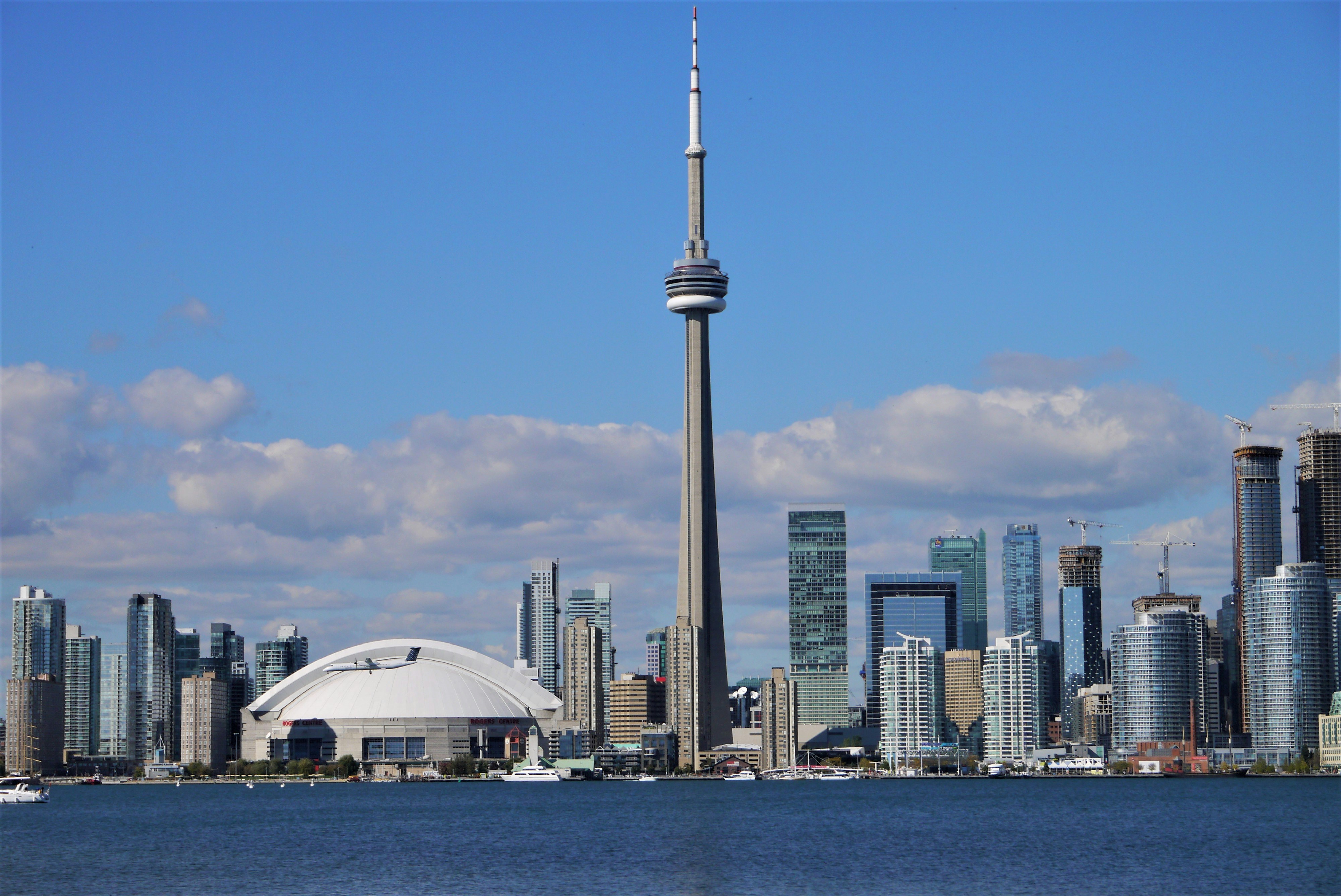 Toronto skyline photograph along the waterfront with the CN Tower rising above glassy high-rises under a clear blue sky. Calm water reflections emphasize modern architecture.