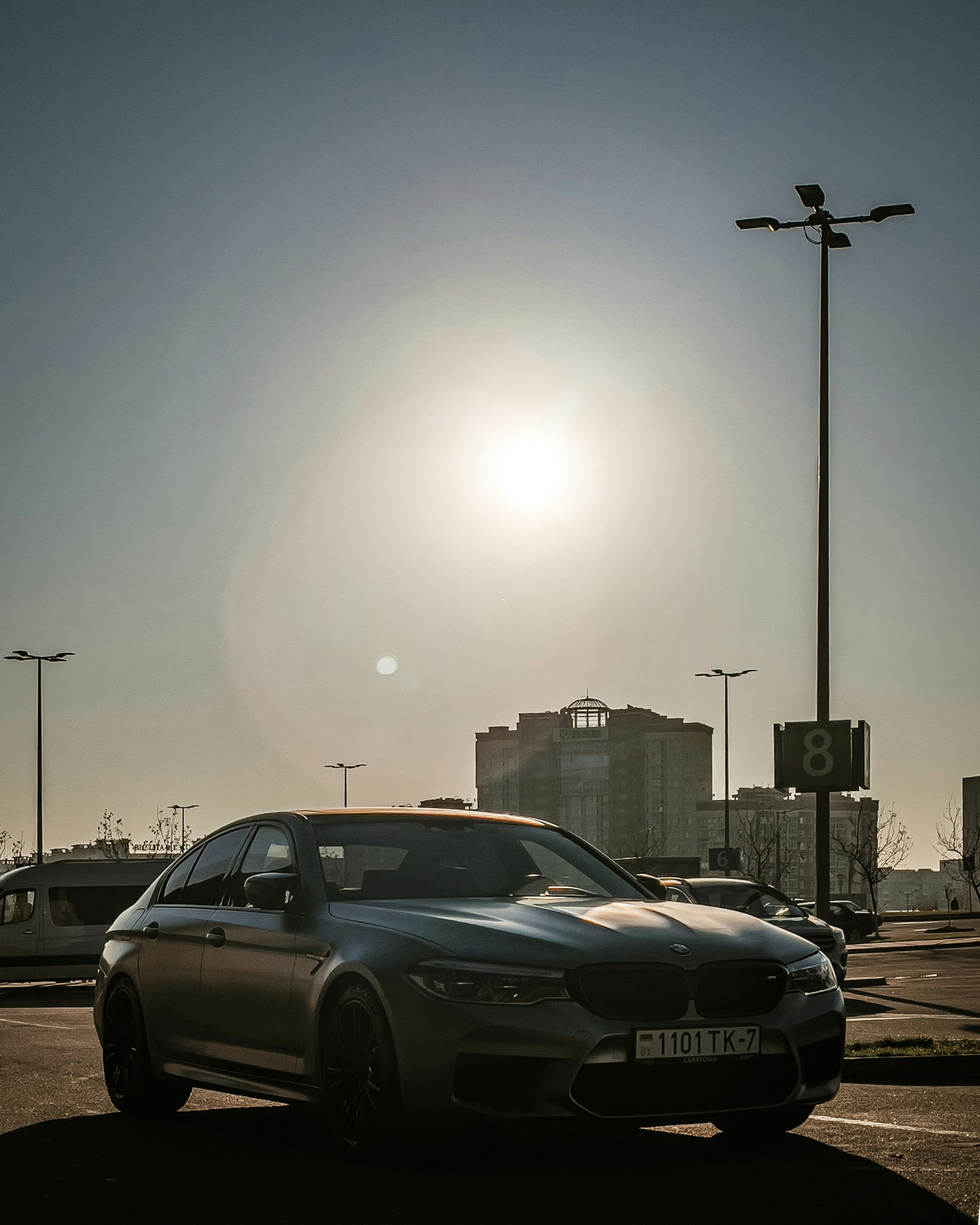 Silver BMW parked in an urban setting under a bright sun, with buildings and parking signs in the background.