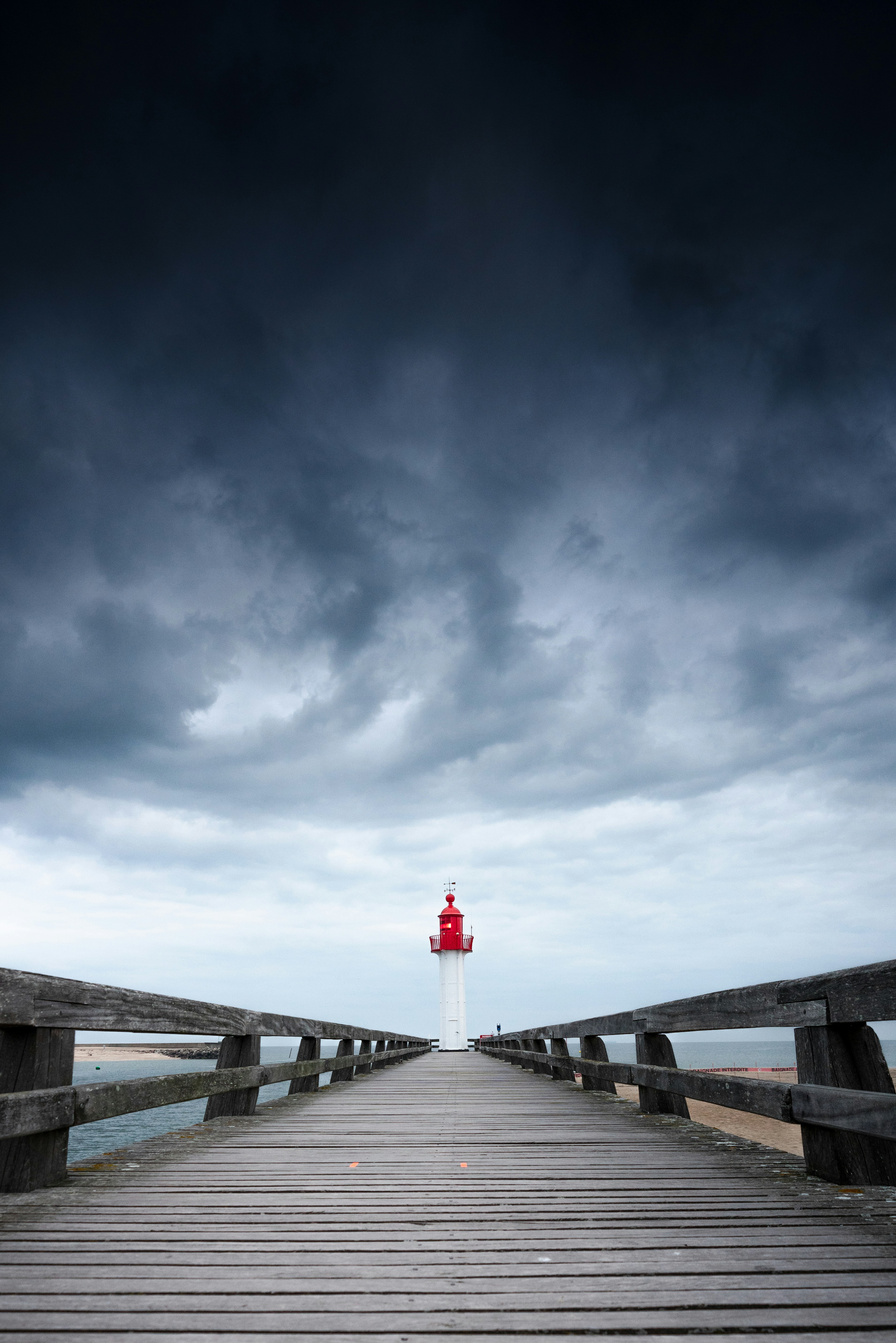 A red and white lighthouse stands at the end of a wooden pier under a dramatic, cloud-filled sky.