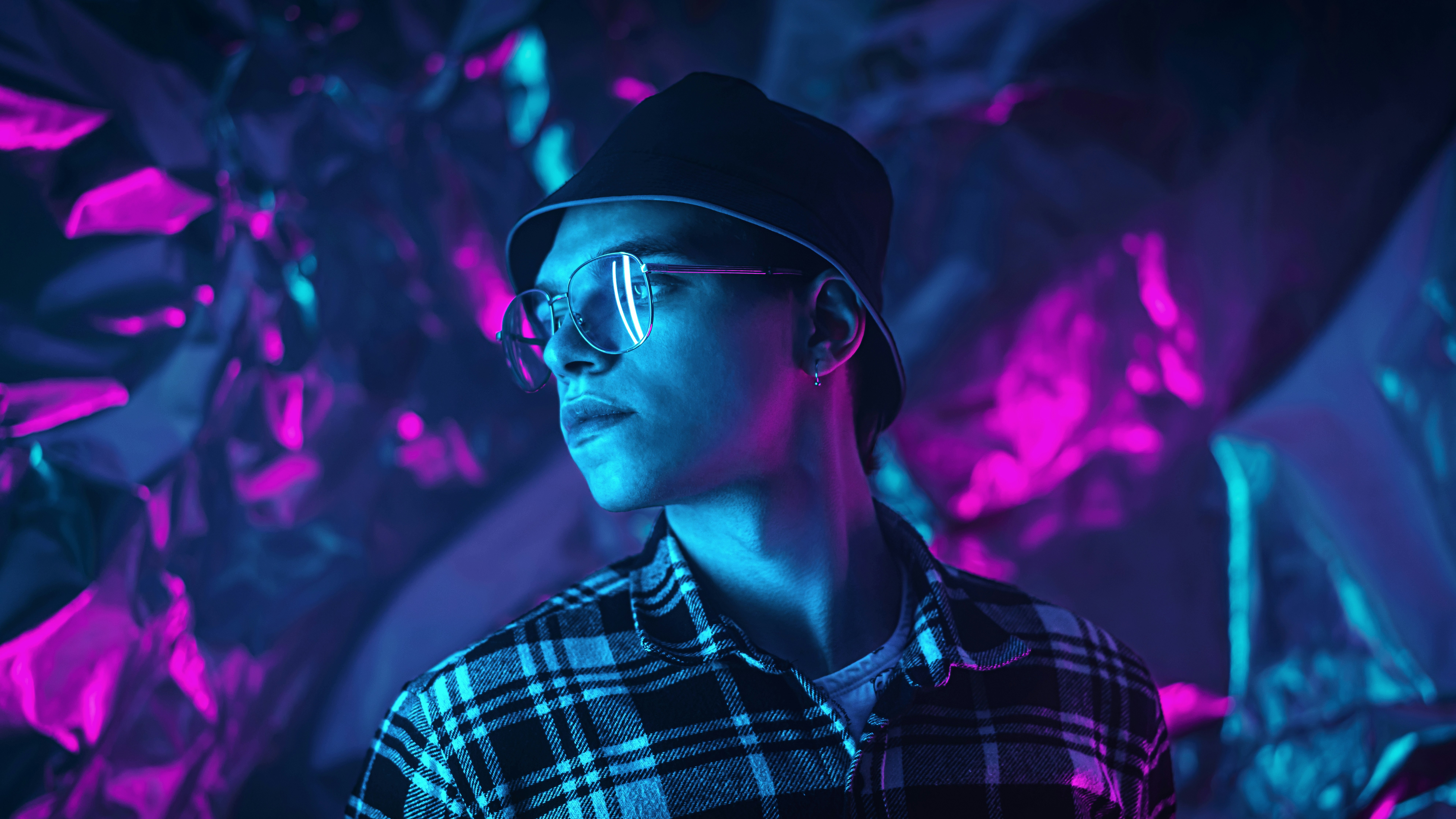 Person in glasses and a hat stands against a metallic backdrop illuminated by neon lights.