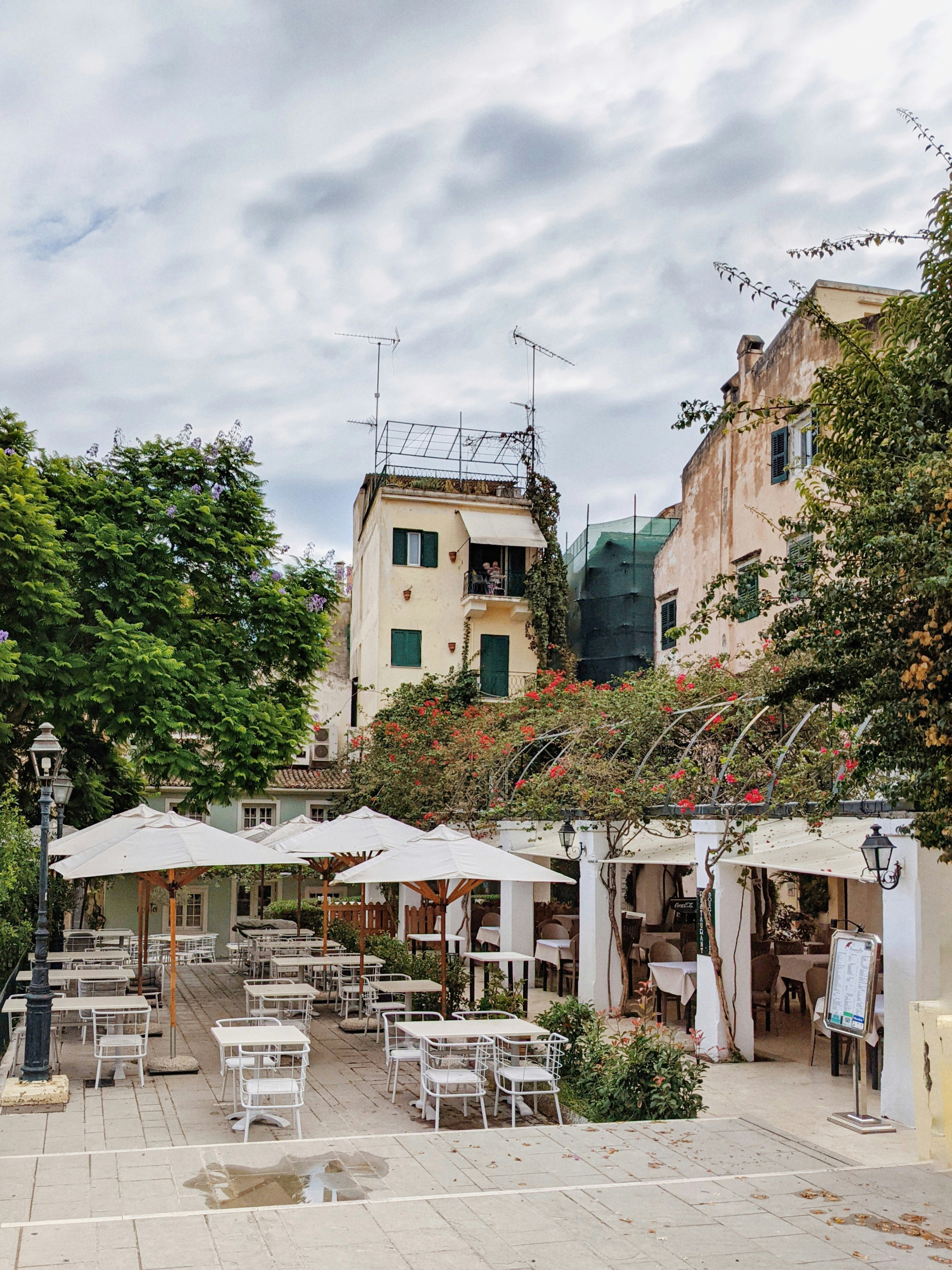 Quaint outdoor dining area featuring white tables and umbrellas surrounded by lush greenery and historic buildings.