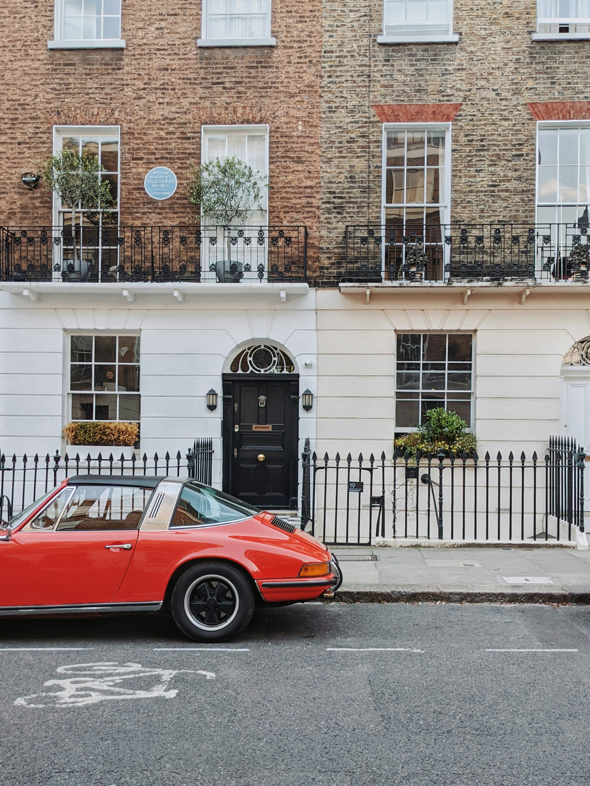 A classic red sports car parked beside a charming Georgian townhouse with decorative window boxes and wrought-iron railings.