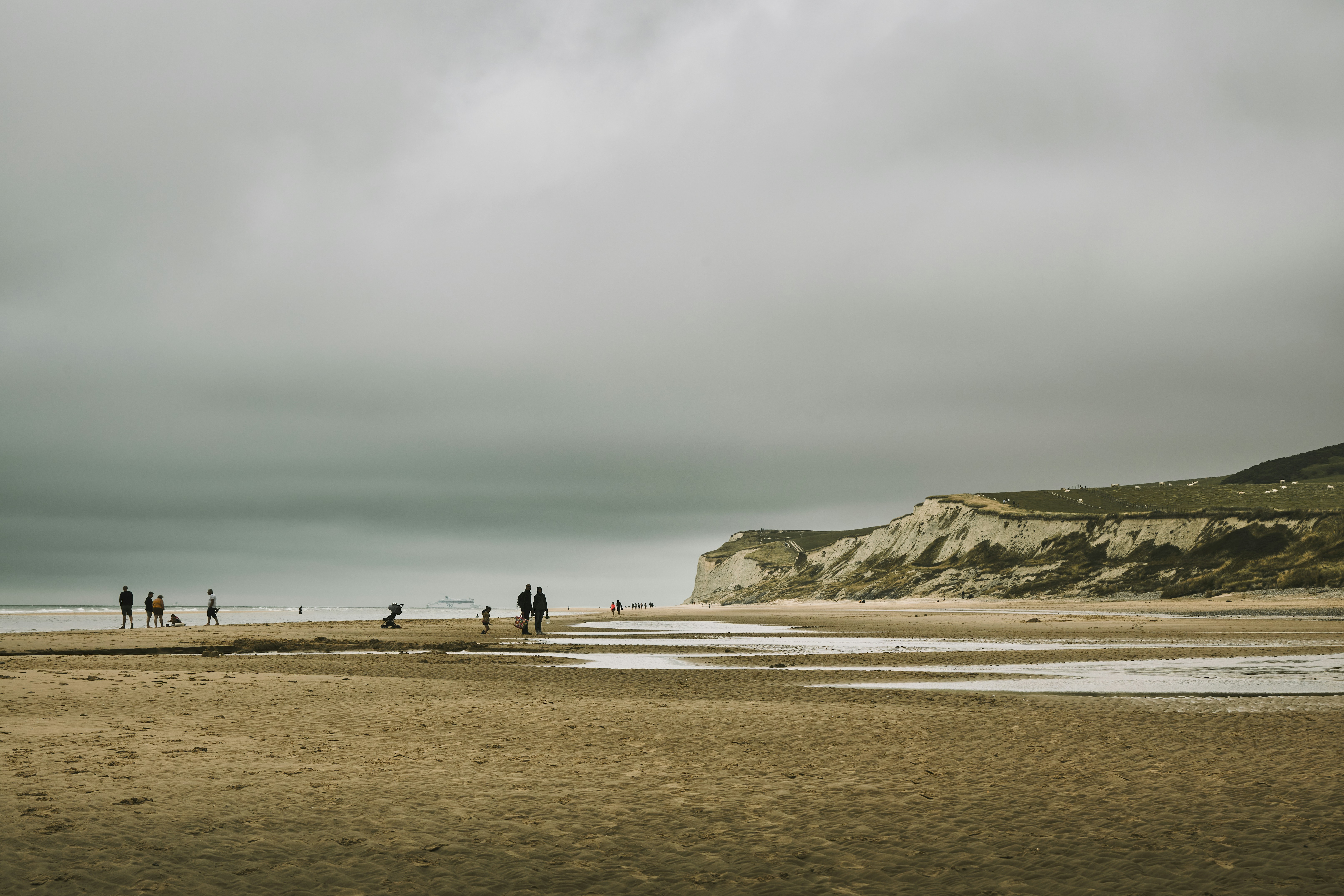 A serene beach scene with silhouettes of people walking along the shore, framed by a rugged cliff under a moody sky.
