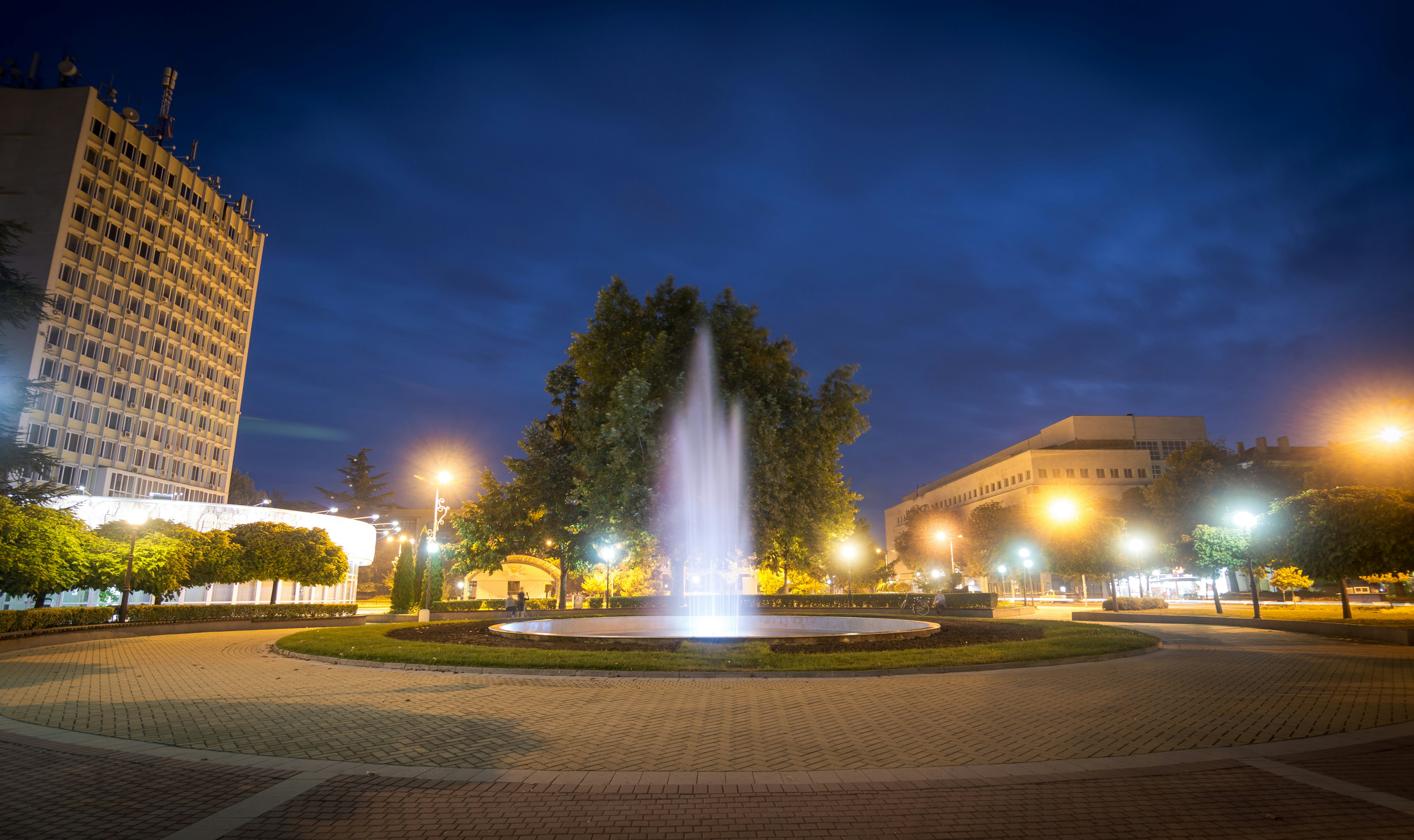 Illuminated fountain surrounded by lush greenery and urban architecture under a twilight sky.