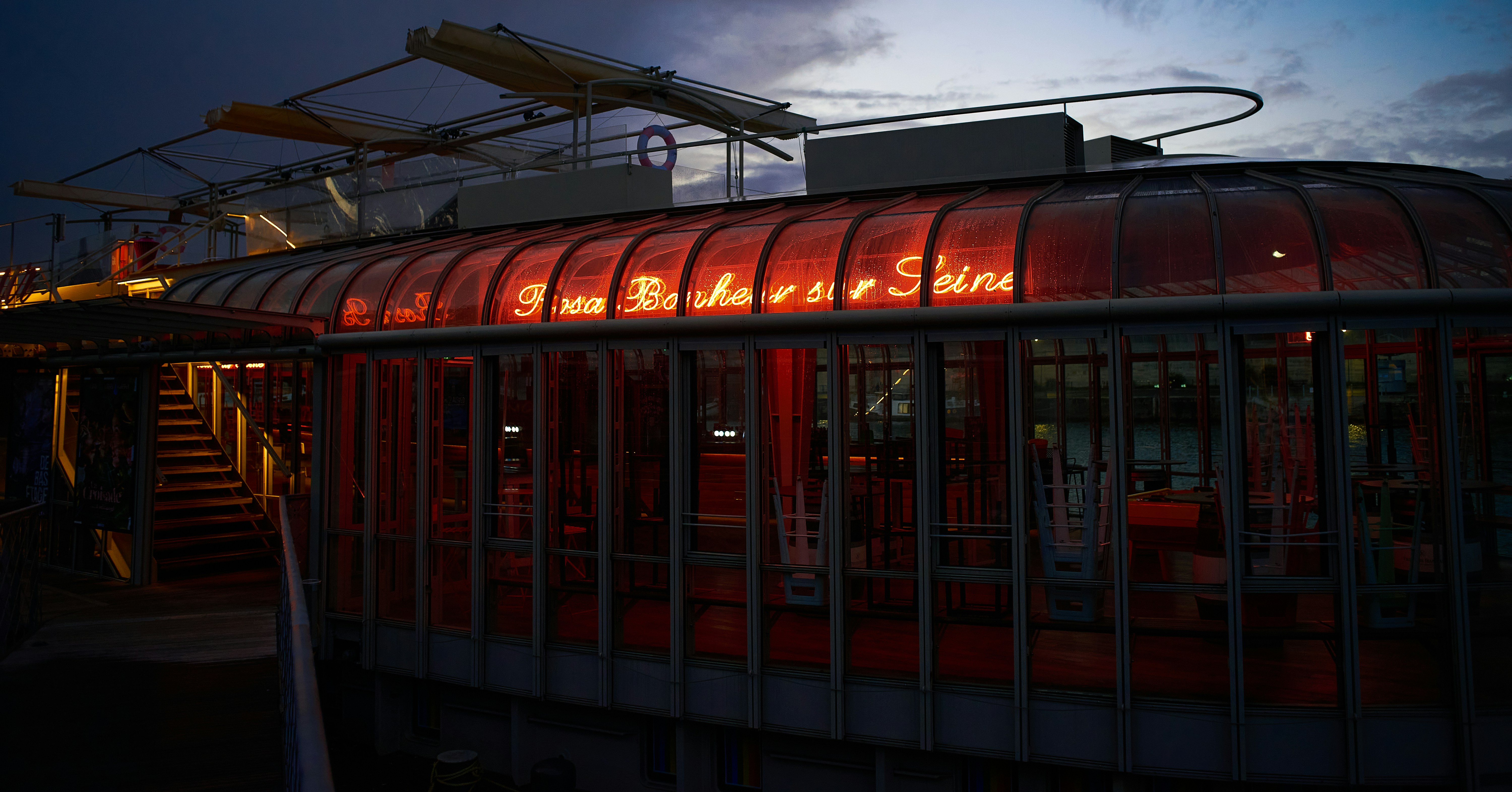 A vibrant restaurant with glowing red signage, nestled on a boat, inviting diners as twilight descends.