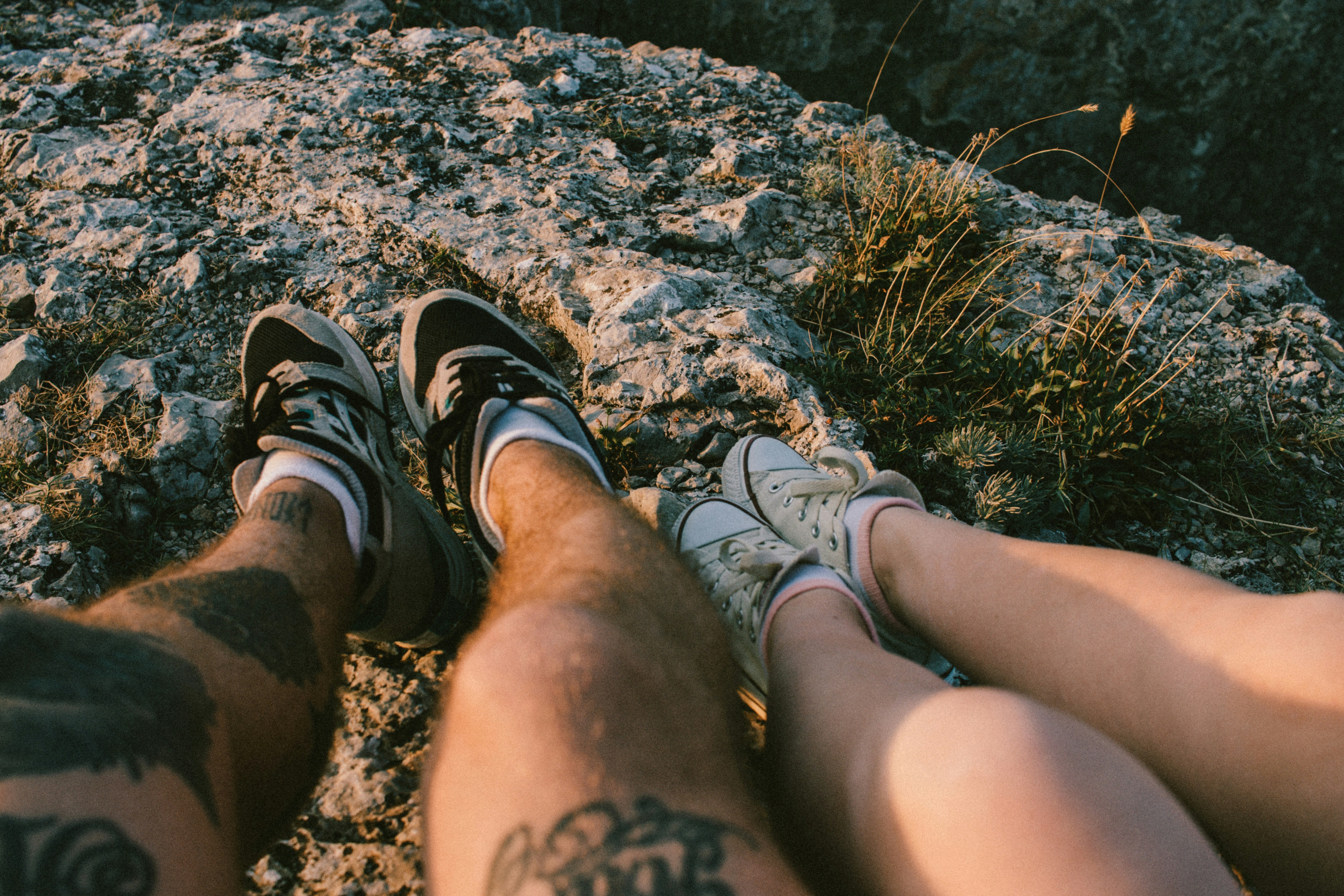 Two pairs of feet resting on a rocky ledge, surrounded by nature, with sunlight casting a warm glow. The scene captures a moment of tranquility and connection.
