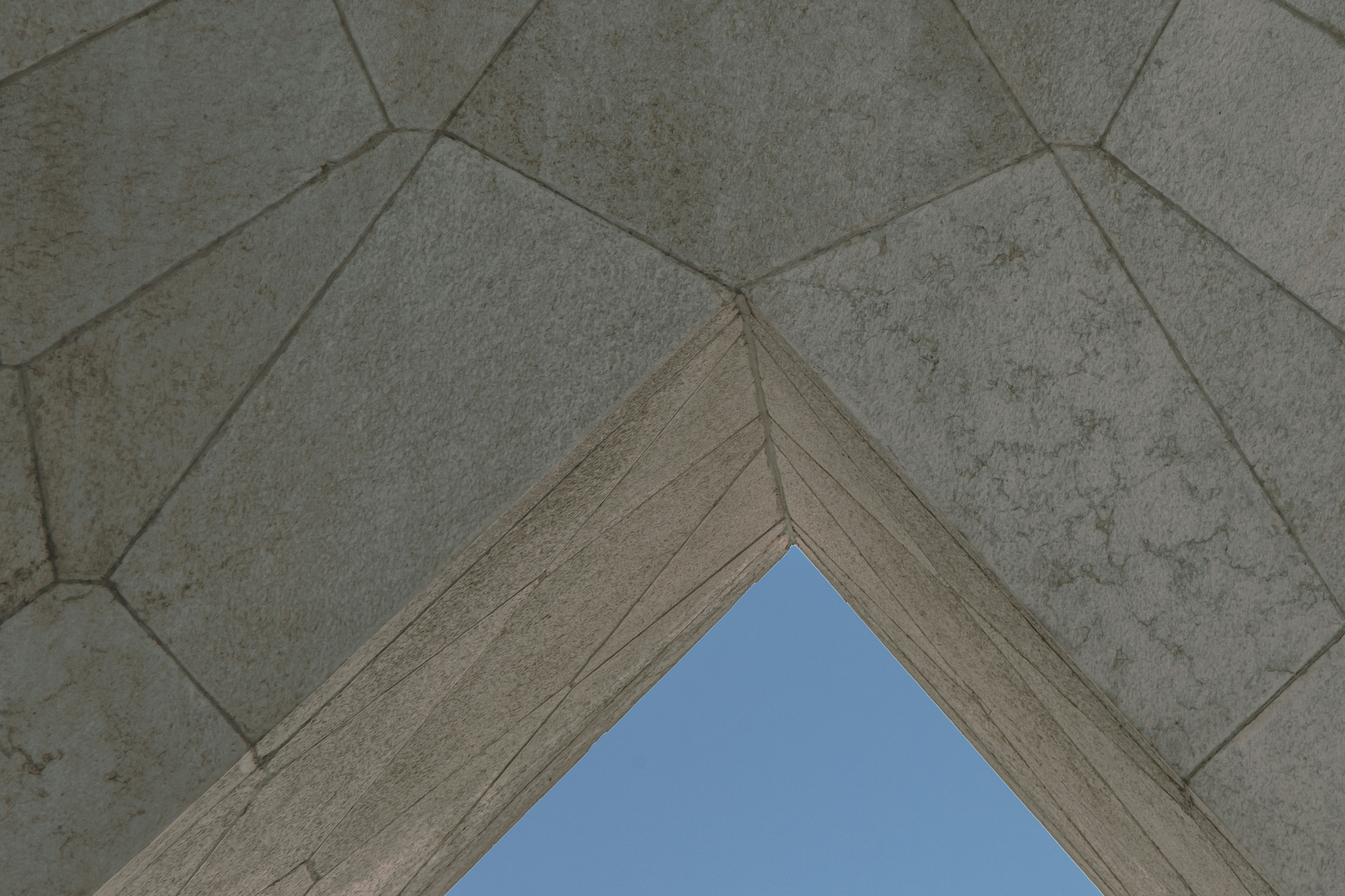 Angular architectural detail revealing a clear blue sky through a triangular opening. The textured surfaces create a striking contrast.