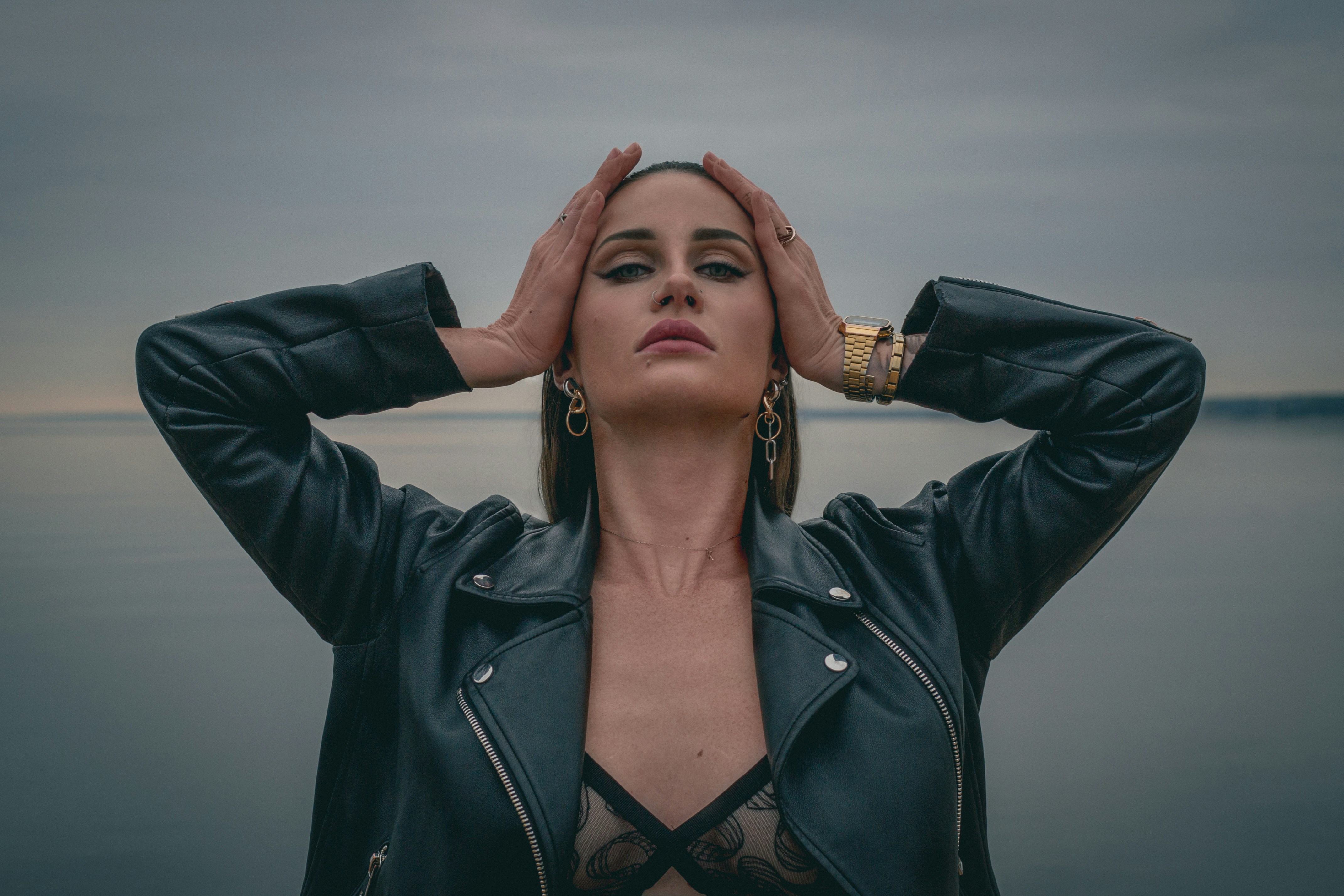 Model with hands on her head, wearing a leather jacket, gazes into the distance against a serene lakeside backdrop.