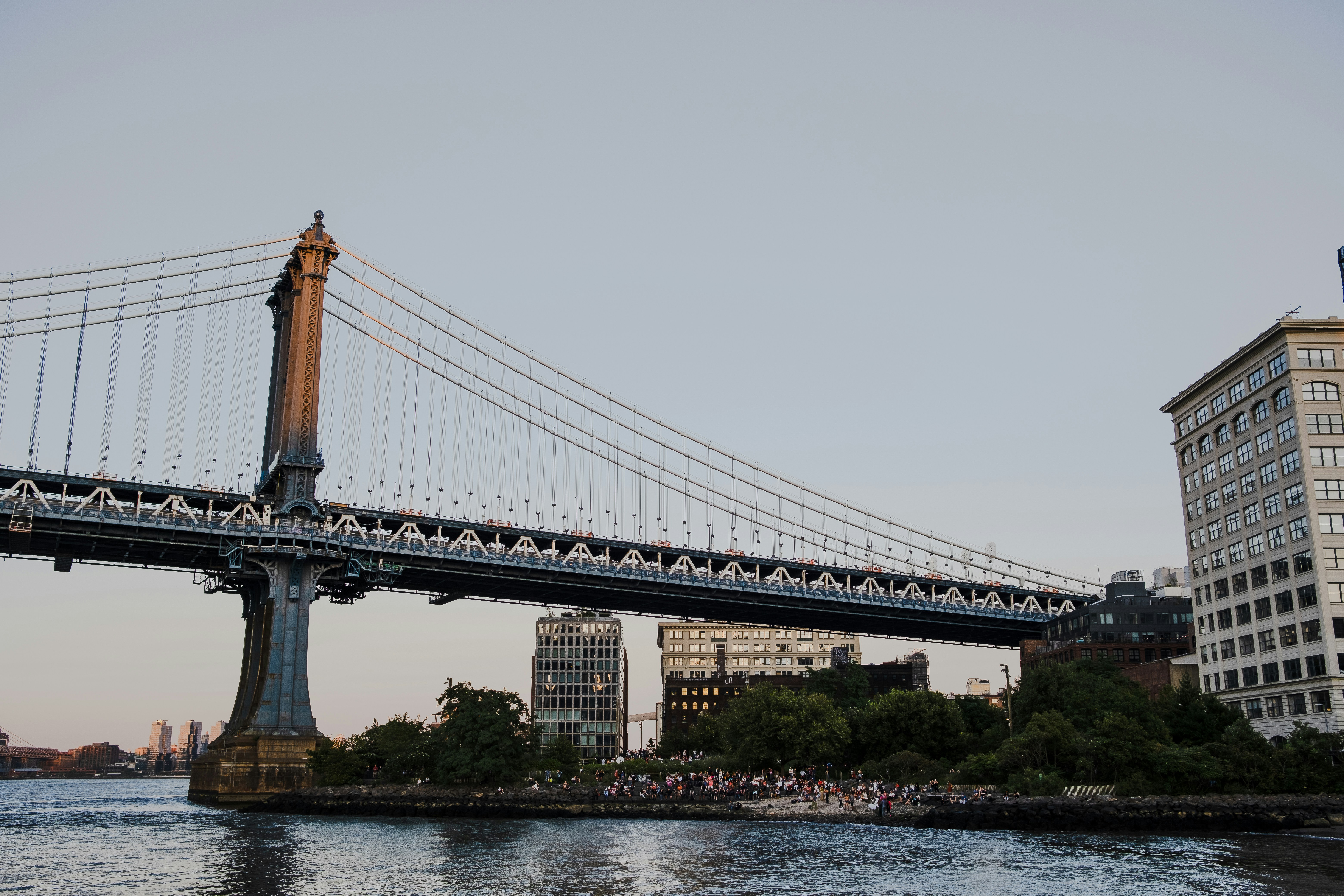 Manhattan Bridge spanning the East River, framed by urban buildings and lush greenery. A vibrant gathering of people is visible along the waterfront.