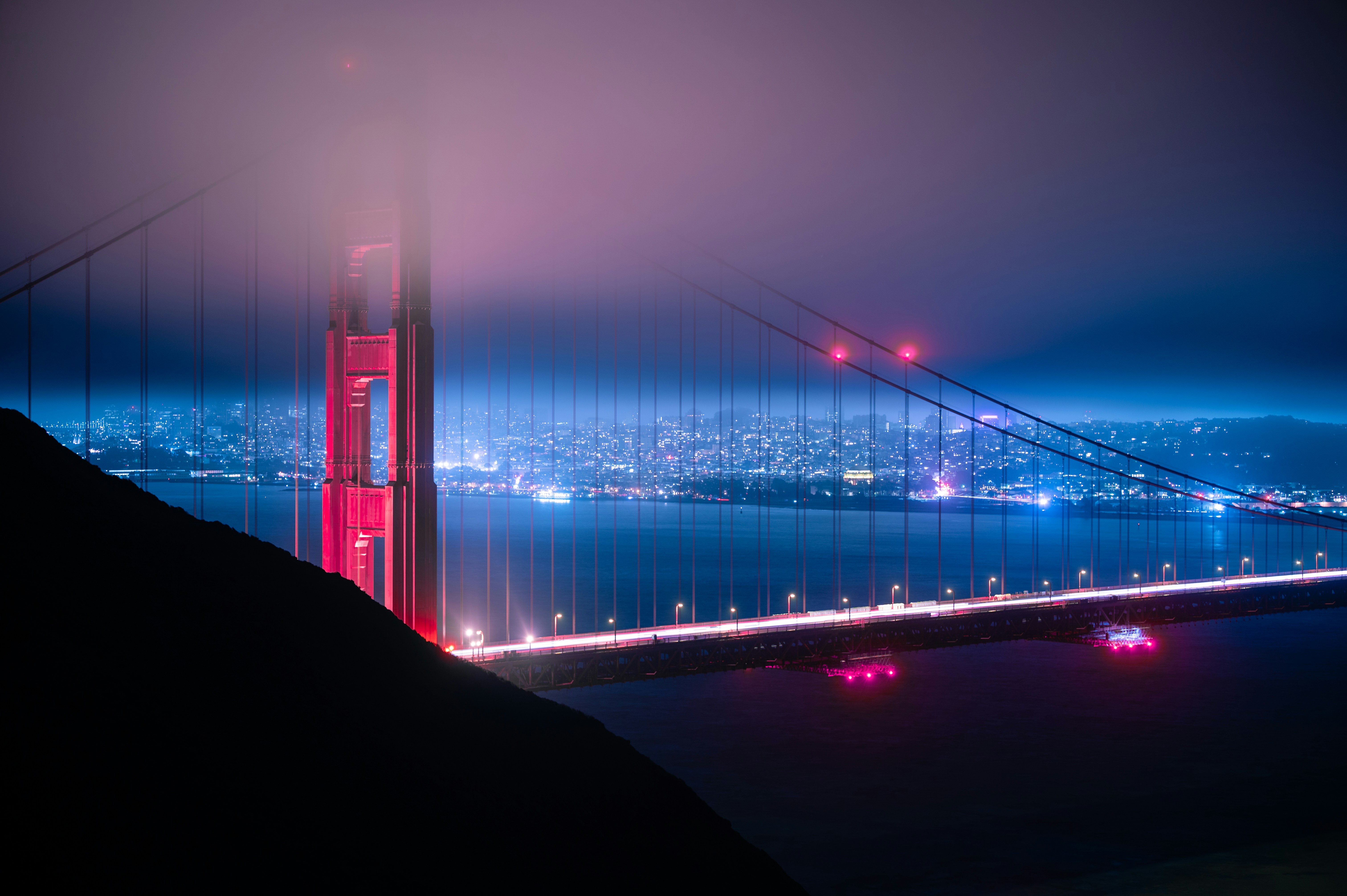 Golden Gate Bridge illuminated in vibrant colors, shrouded in fog with a city skyline in the background. A striking display of urban beauty at night.