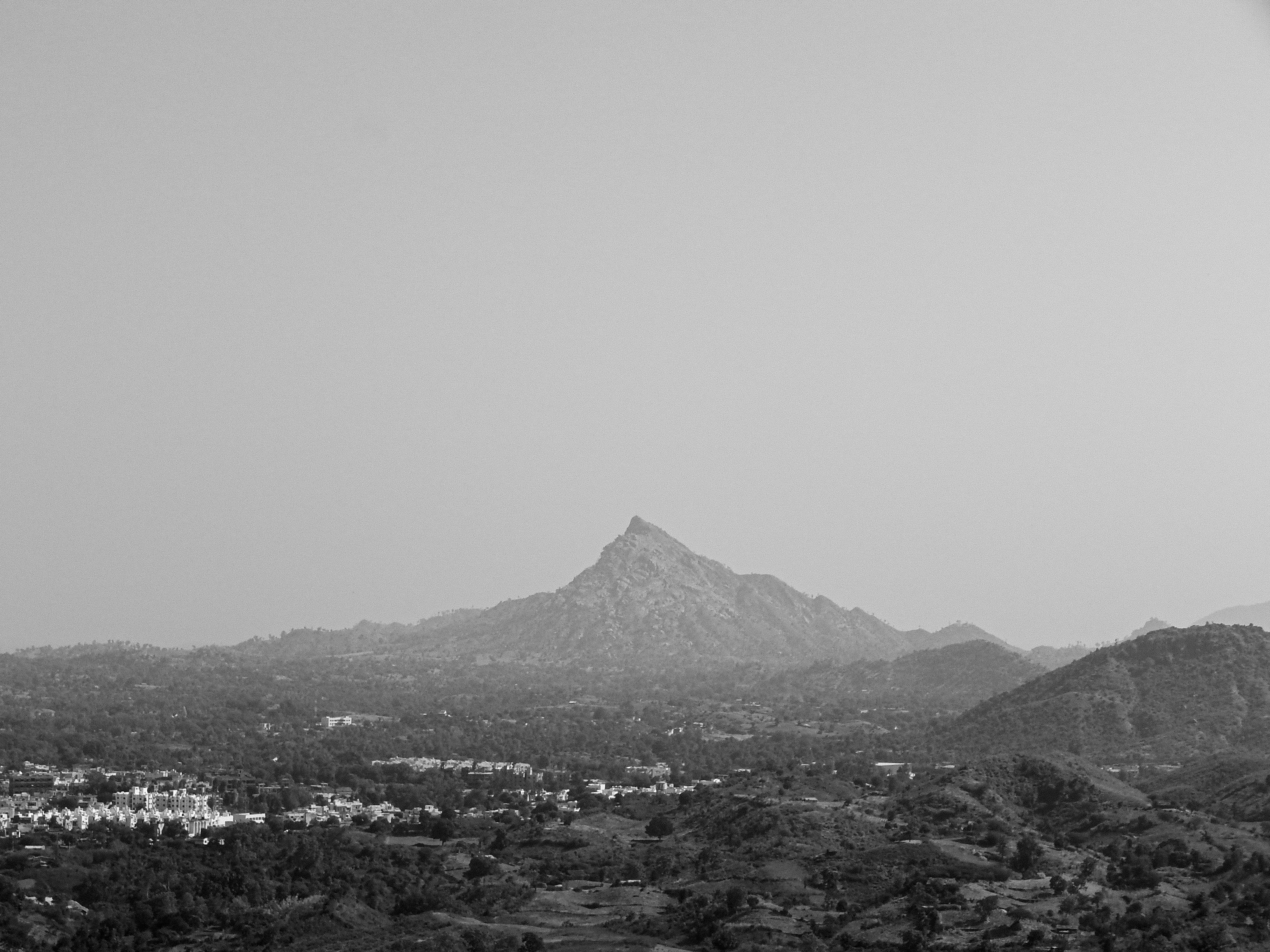 A monochromatic landscape featuring a prominent mountain peak rising over a sprawling valley, with distant hills and a small town visible below.