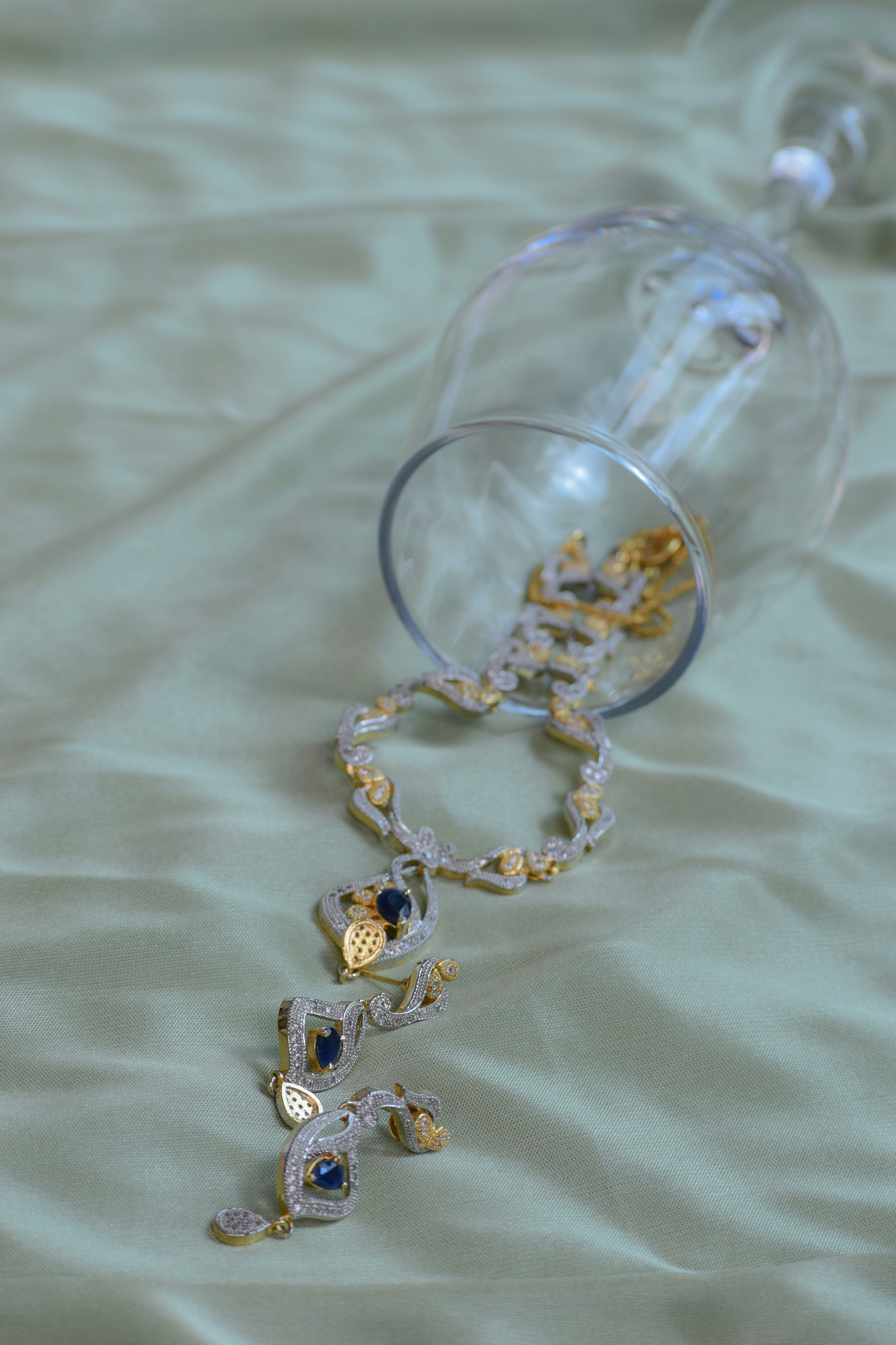 A delicate gold and silver bracelet cascades from an overturned glass onto a soft green fabric, showcasing intricate design and craftsmanship.