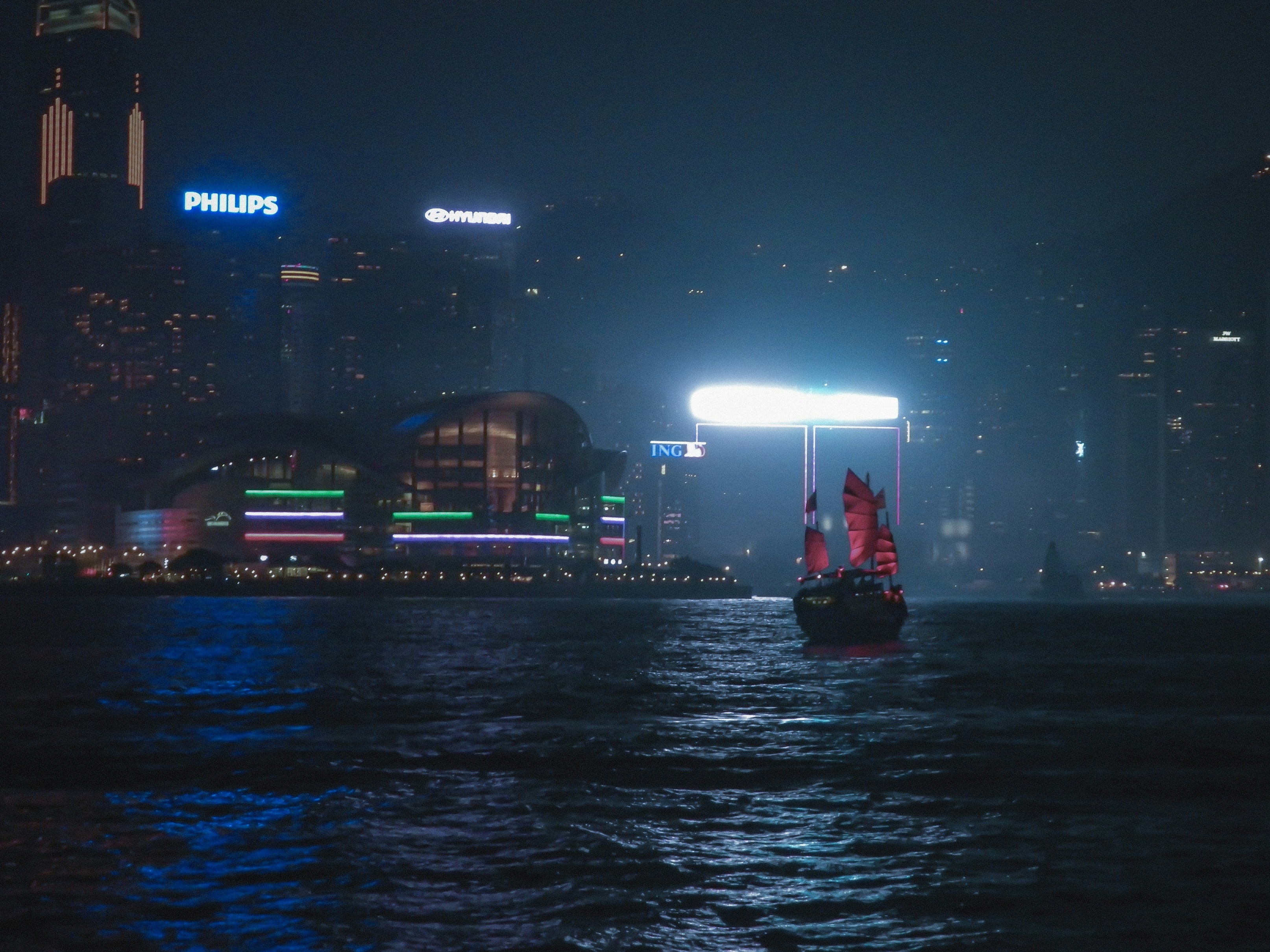 Traditional boat with red sails gliding through a city harbor at night, surrounded by glowing skyscrapers.