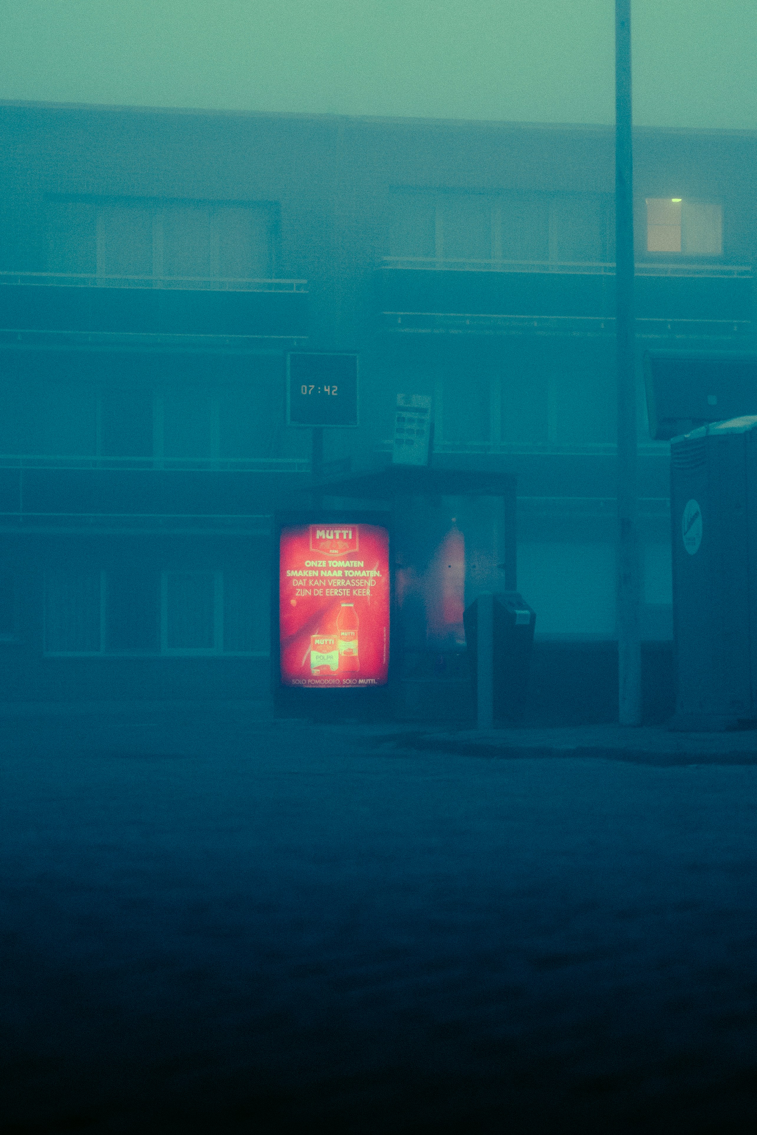 A bus stop illuminated by a bright advertisement stands in a foggy urban landscape, evoking a sense of solitude and mystery. The soft hues of the scene create an ethereal atmosphere.