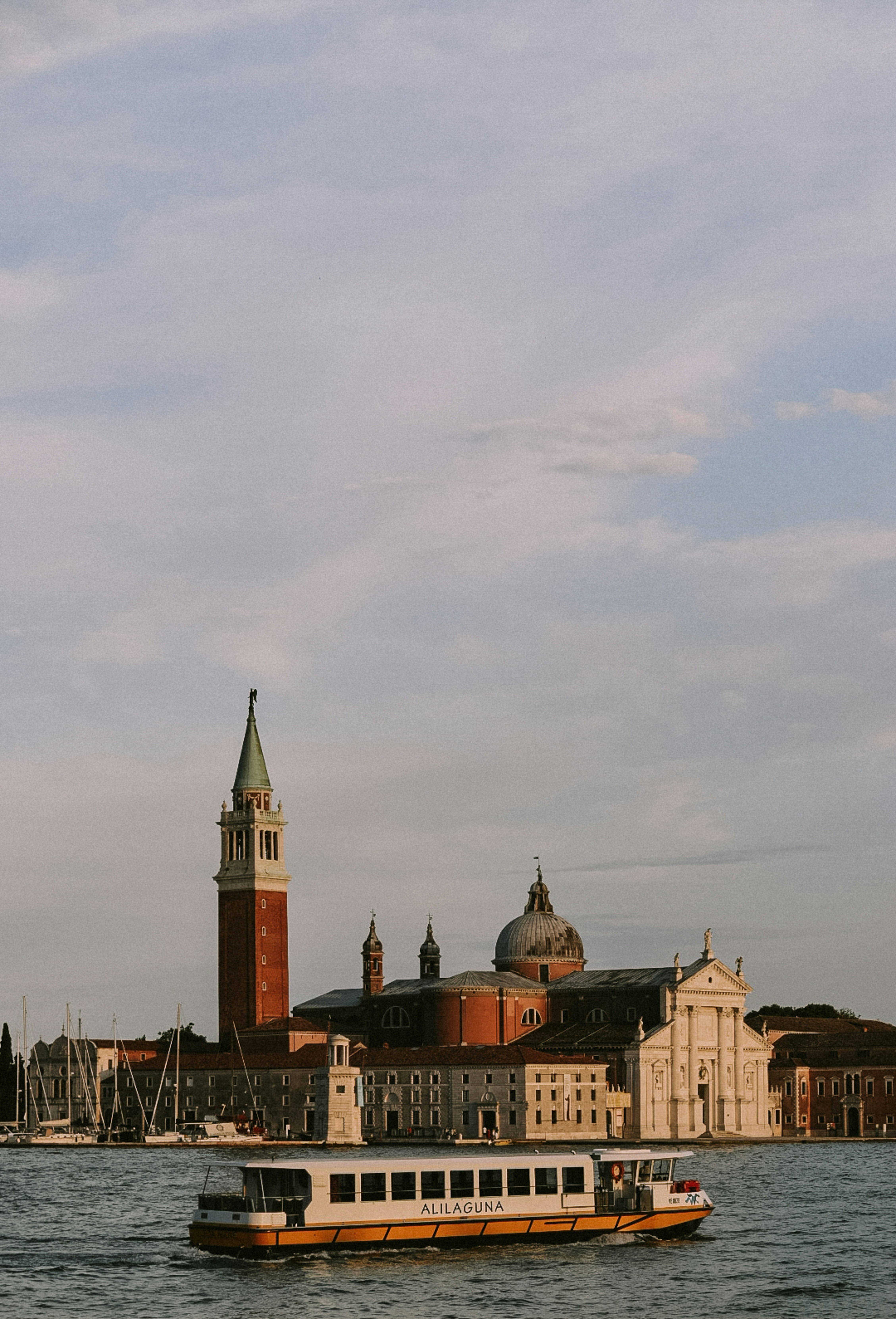 A boat labeled 'ALILAGUNA' glides across the tranquil waters of Venice, framed by historic architecture and a soft sky.