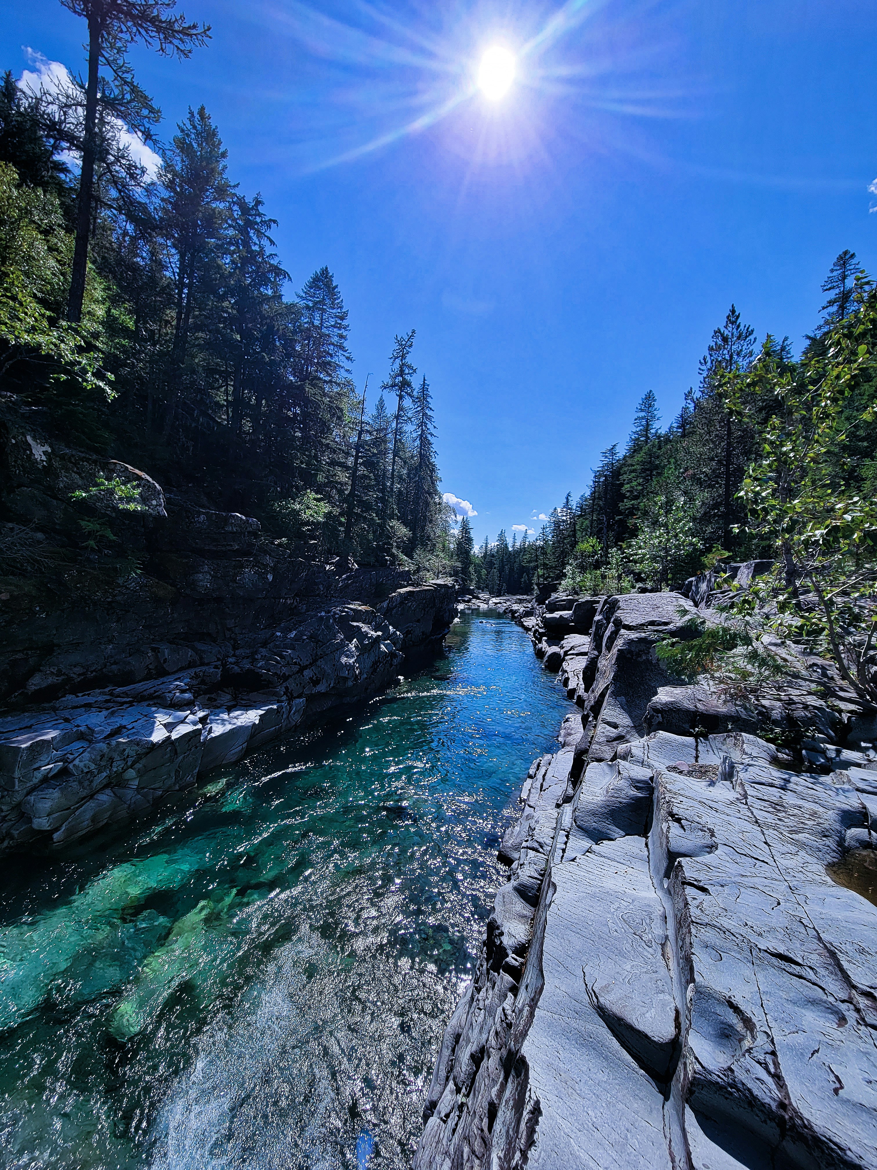 A serene river flows between rocky banks, surrounded by lush trees under a clear blue sky. Sunlight glimmers on the water's surface.