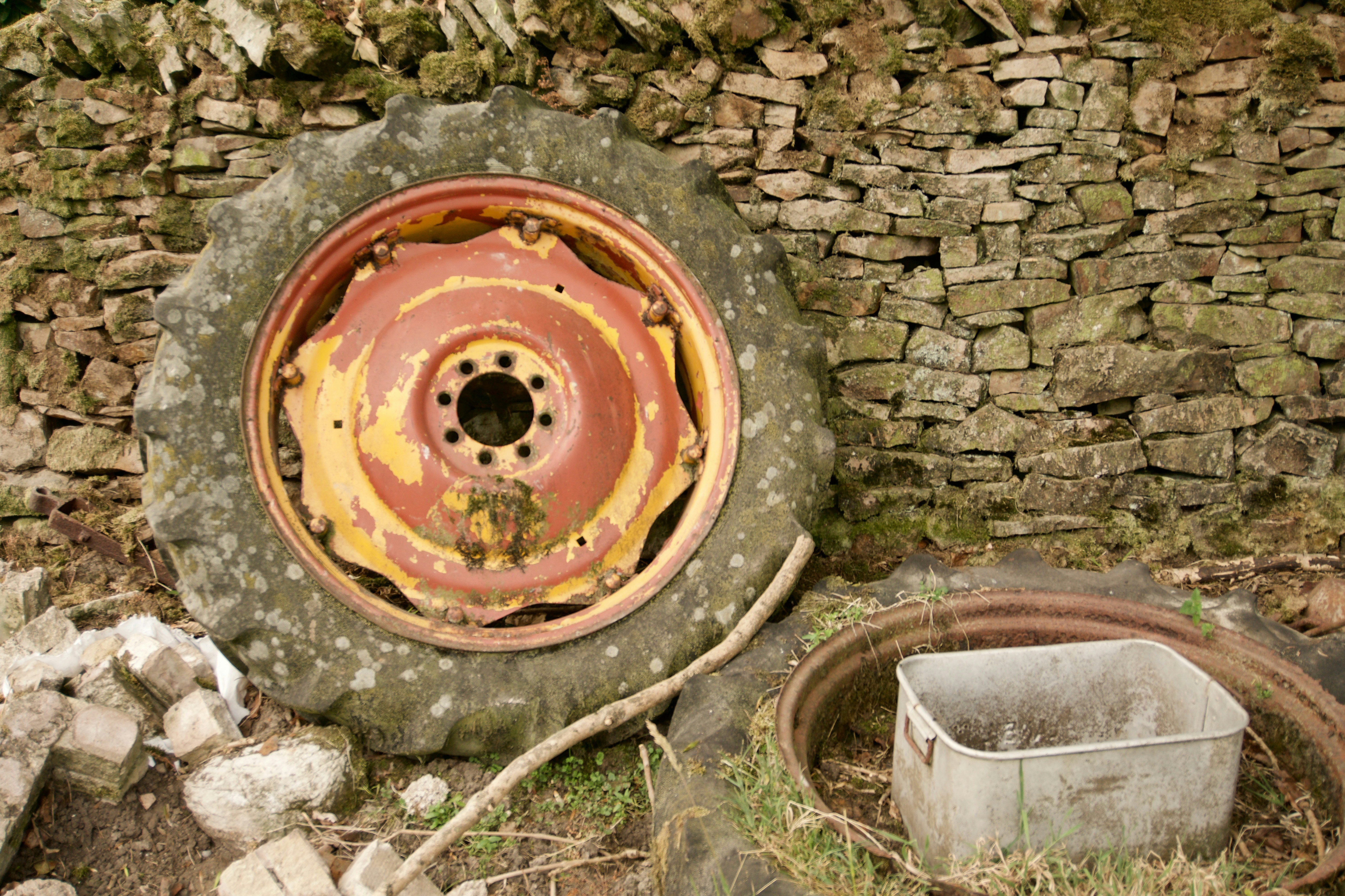 A weathered tractor wheel, painted in faded yellow and red, leans against a moss-covered stone wall, creating a captivating juxtaposition of textures and colors. The muted tones and soft natural light evoke a sense of quiet decay and rural nostalgia, offering a visually rich image perfect for design projects needing a touch of rustic charm. The composition's balance between organic and man-made elements adds depth and visual interest.