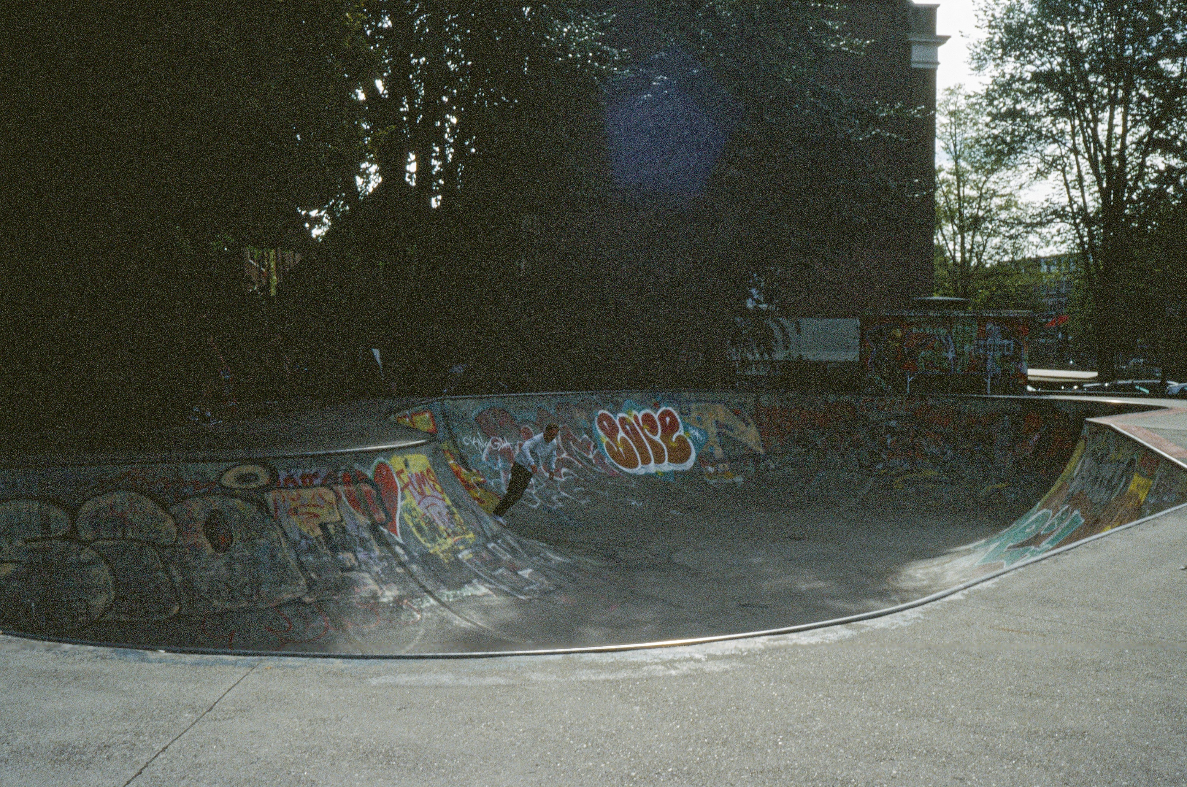 Photograph of a graffiti-painted skate bowl in a quiet park, with a lone skater along the edge and mural-covered walls in the background.