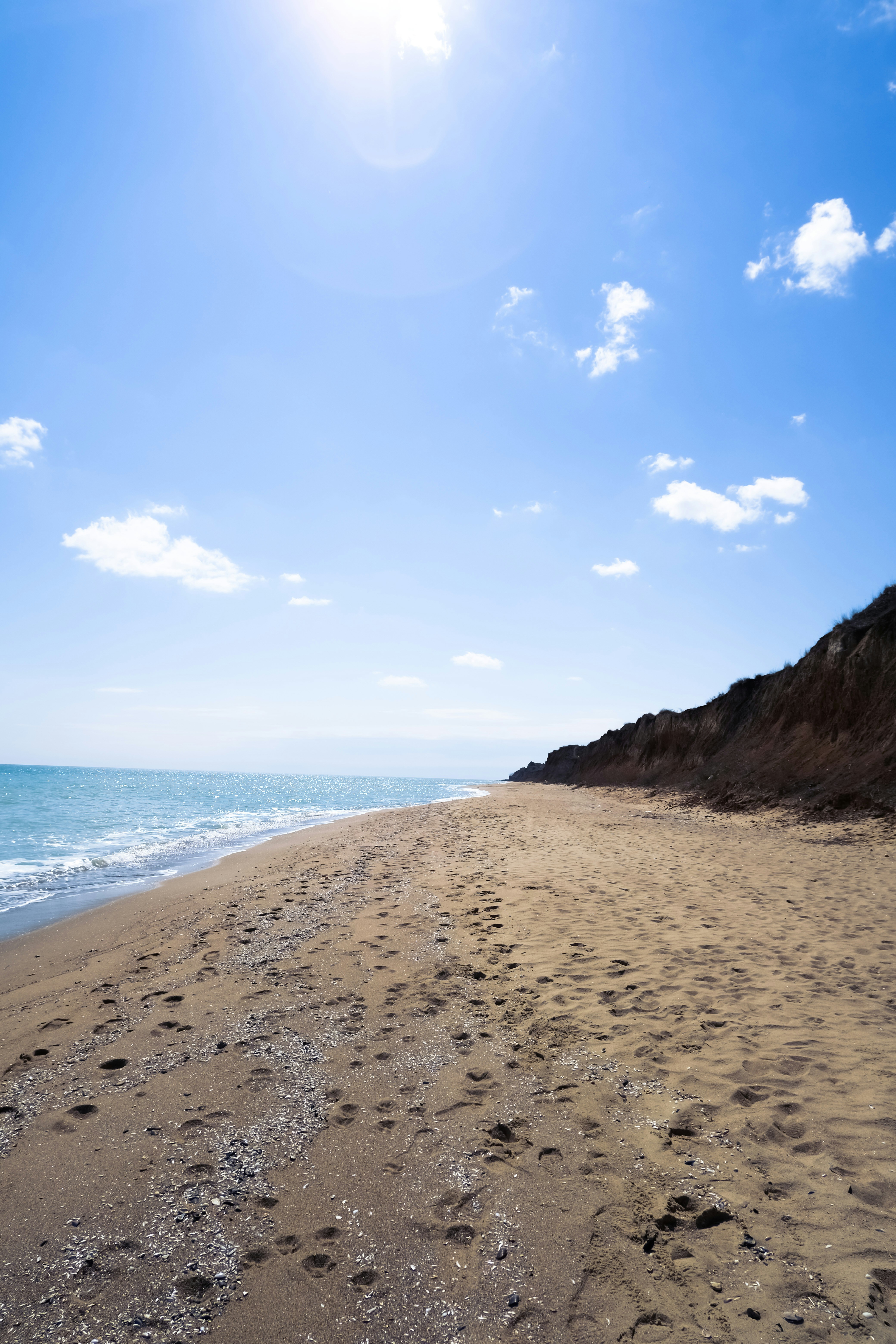 A tranquil beach scene featuring footprints in the sand leading toward a calm ocean under a bright blue sky with scattered clouds.