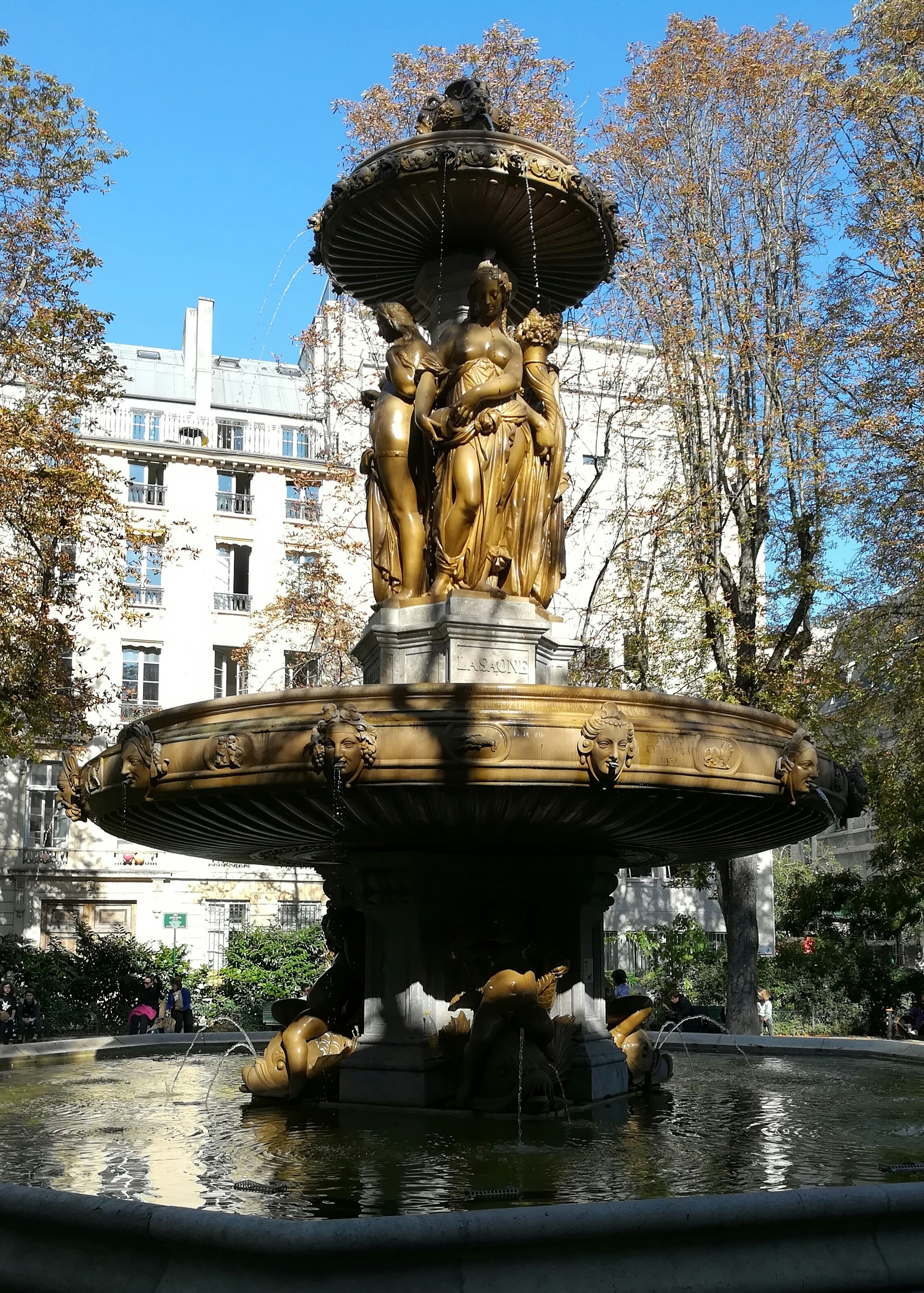 Bronze multi-tier fountain with sculpted figures stands in a leafy city square beneath a bright blue sky.