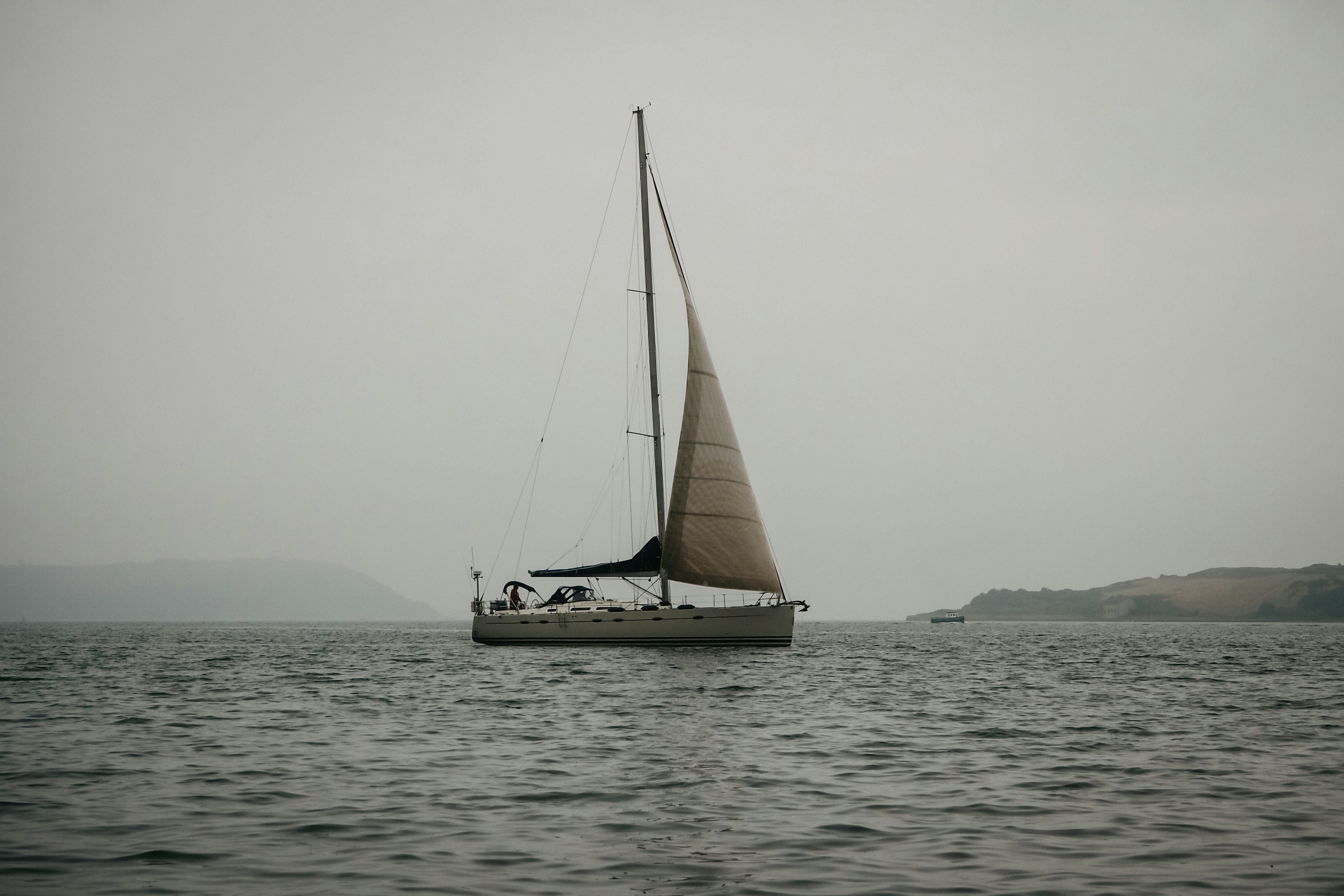 Sailboat gliding across a calm, foggy sea, with a distant shoreline barely visible. The scene evokes a sense of tranquility and adventure.