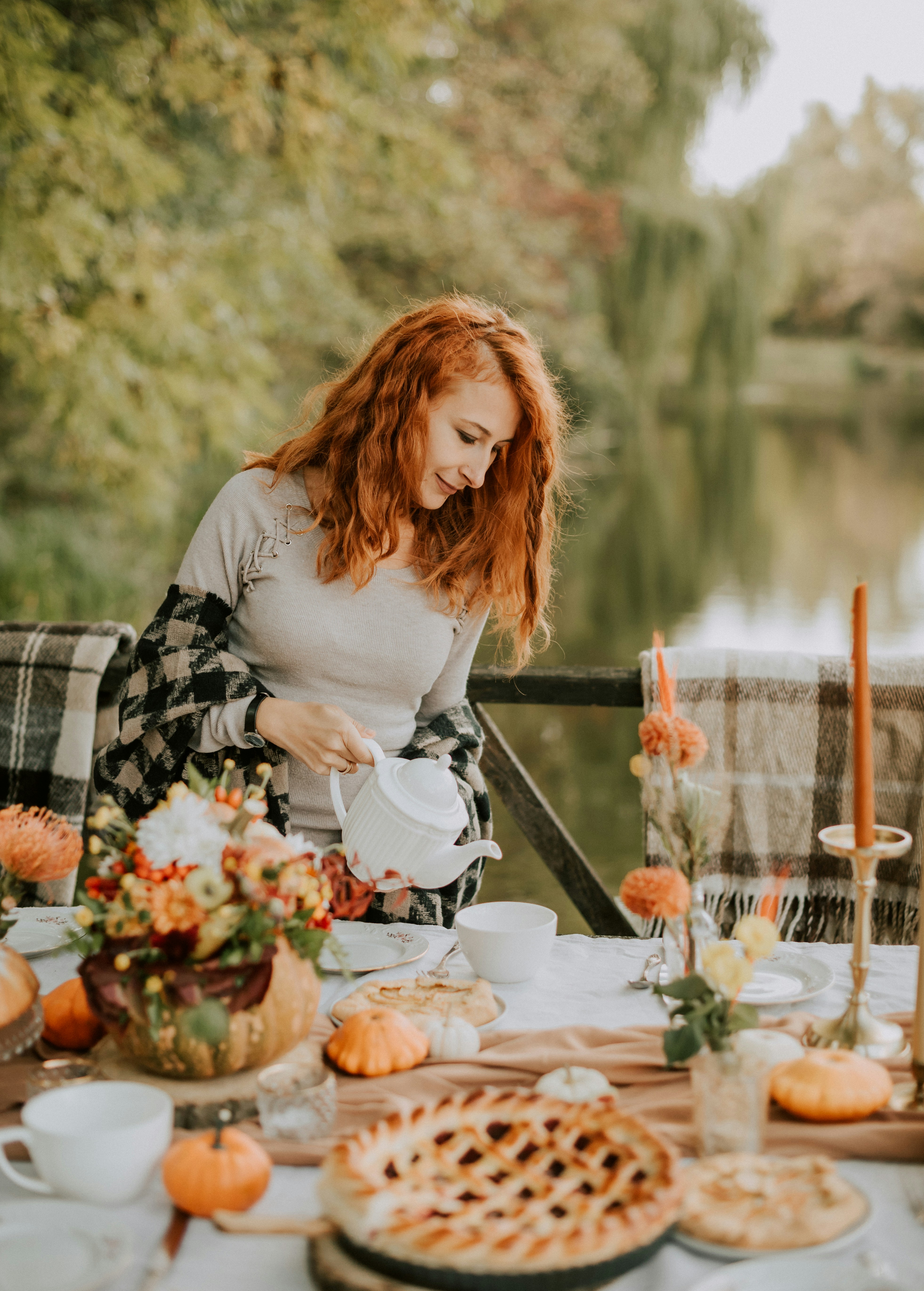 Autumn lakeside tea scene featuring a woman with red hair pouring from a kettle at a long table laid with pies, pumpkins, flowers, and candles. This photograph captures a cozy, sun-washed moment of hospitality by the water.
