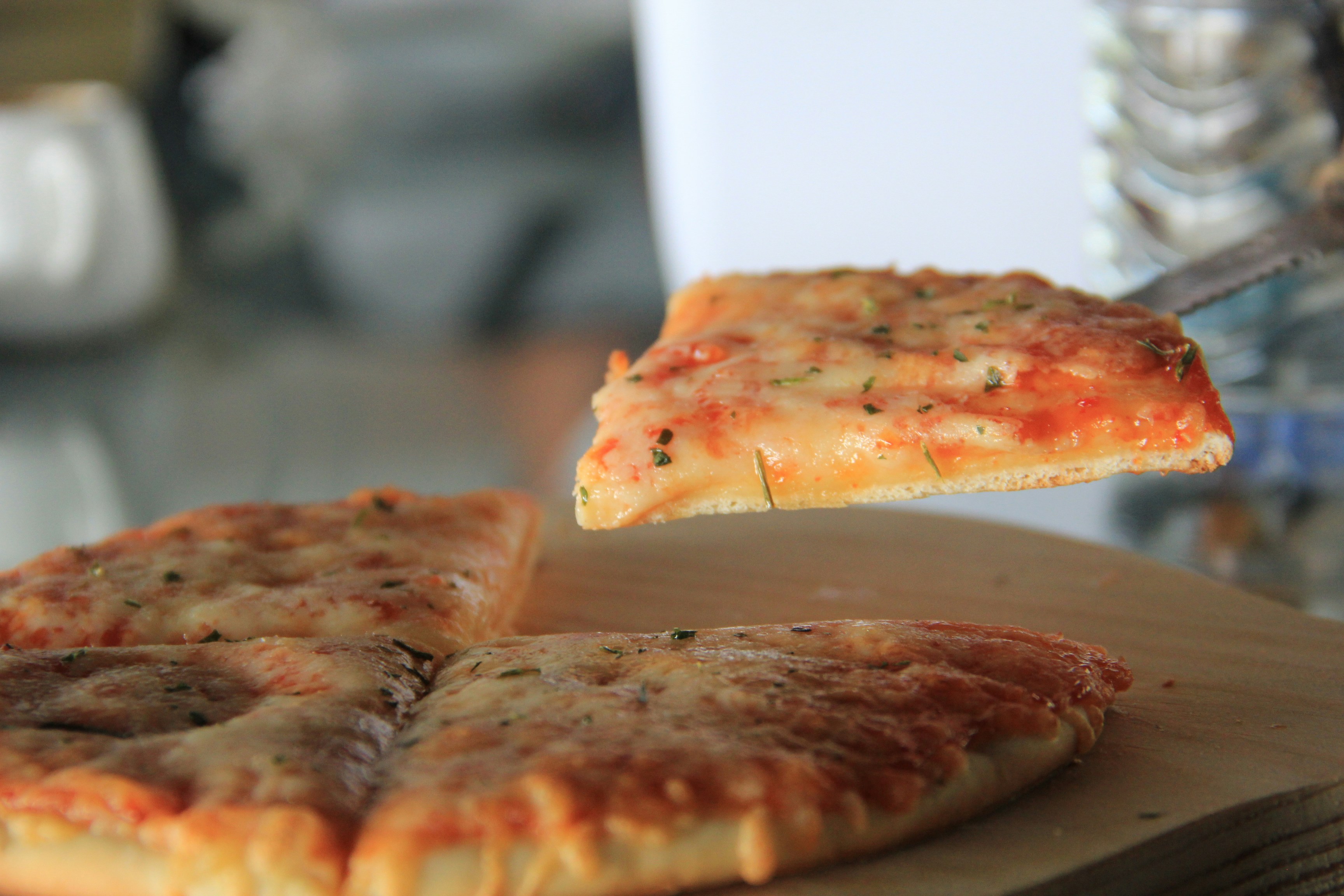 Cheesy pizza slice being lifted from a wooden board, showcasing melted cheese and herbs. The background features a blurred setting, emphasizing the deliciousness.