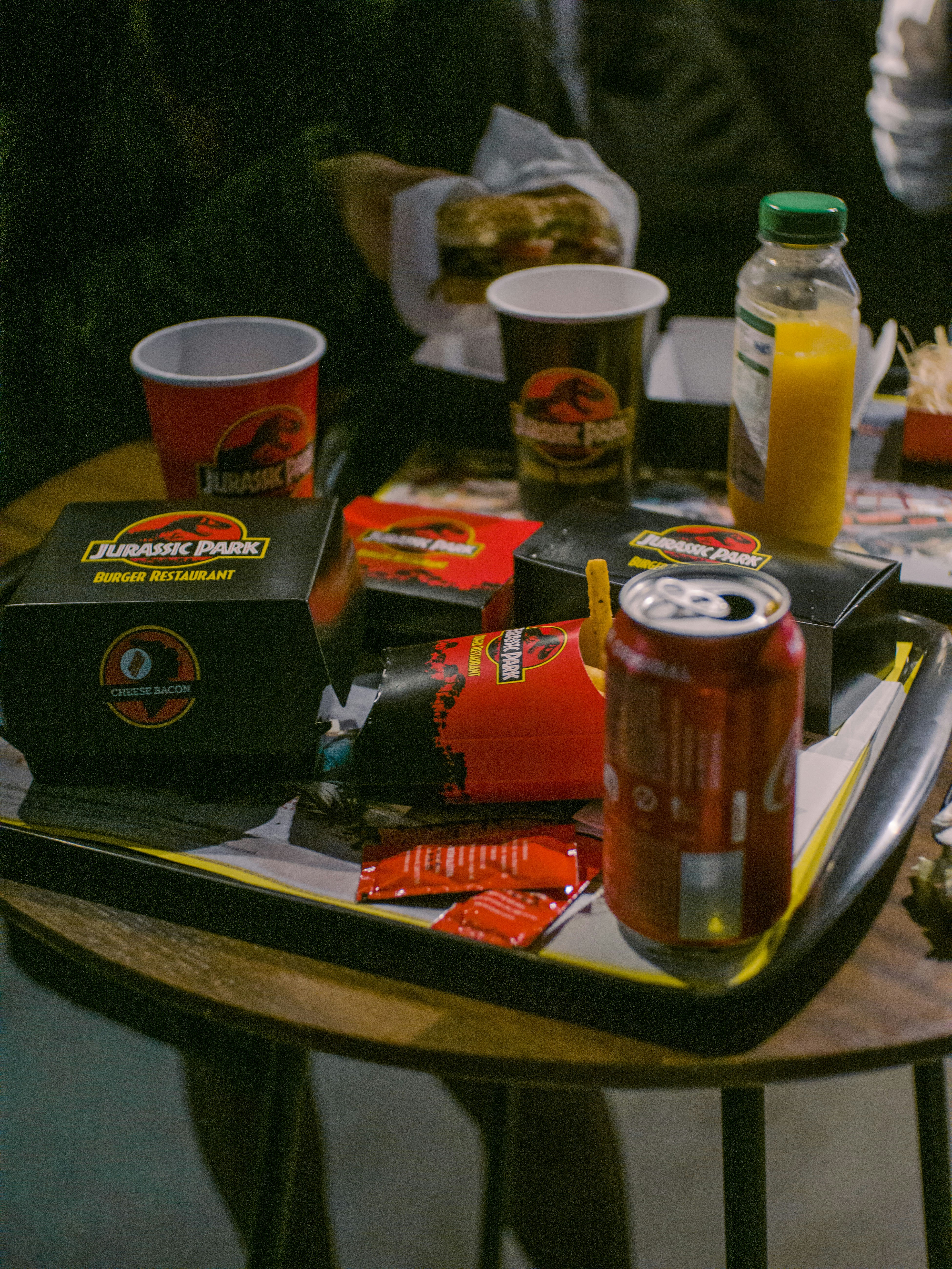 A vibrant display of Jurassic Park-themed food items on a table, featuring burgers, drinks, and condiments. The atmosphere hints at a playful dining experience inspired by the iconic franchise.