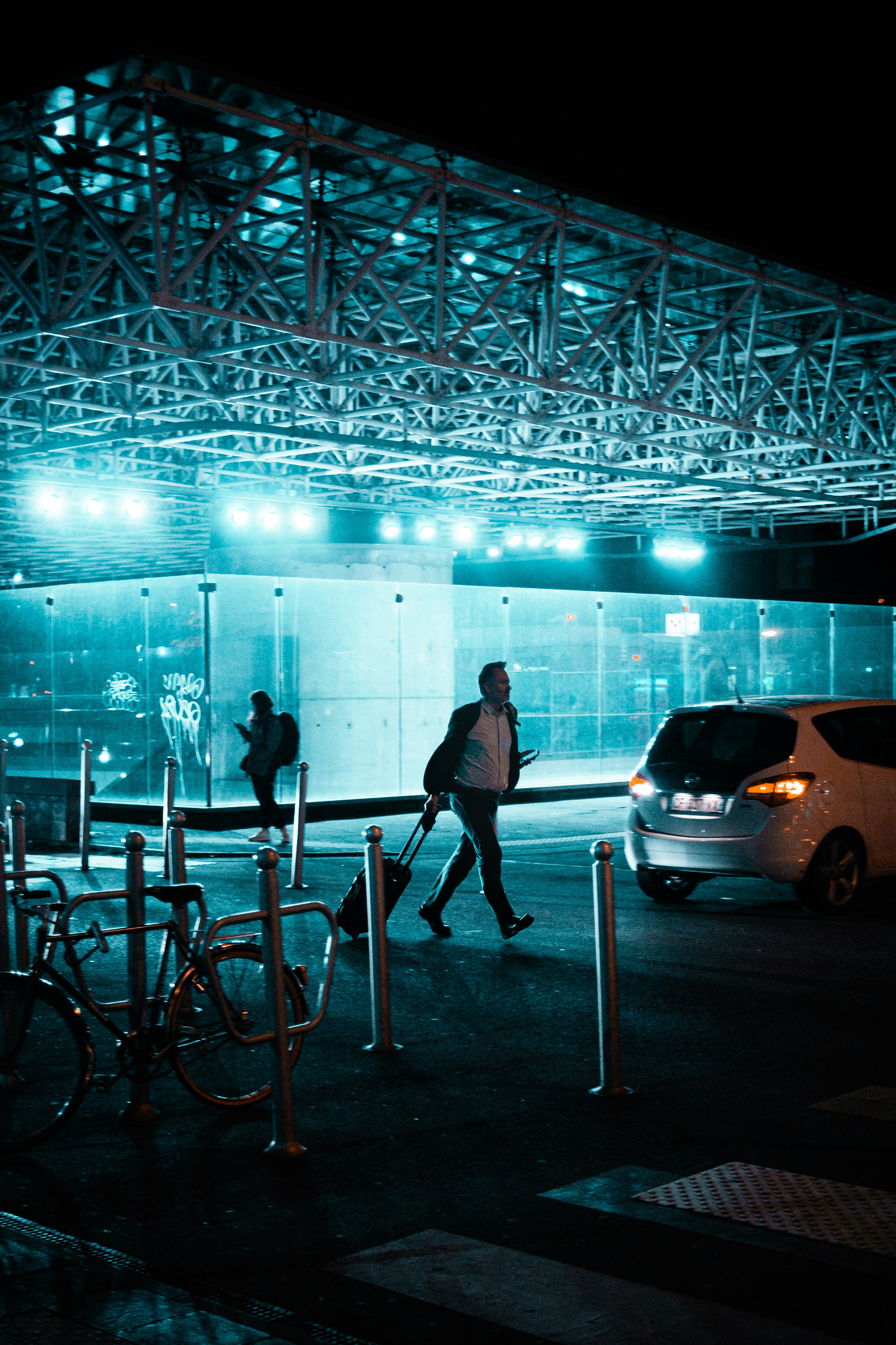 A man walks with a suitcase in a vibrant urban setting illuminated by blue lights, while a car passes by and bicycles are parked nearby.