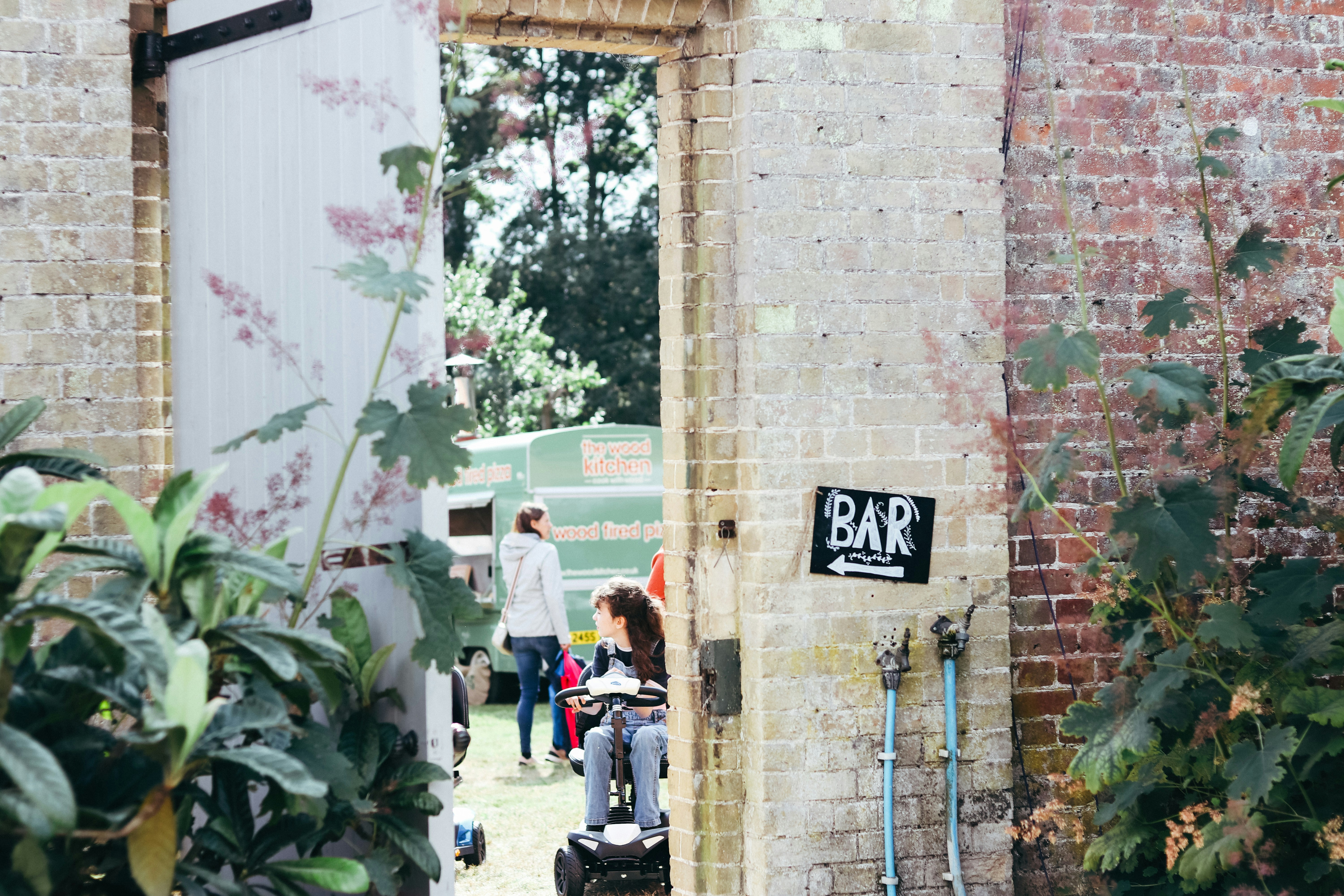Open doorway revealing a lively outdoor scene with people enjoying a bar area, framed by greenery and rustic brick walls.