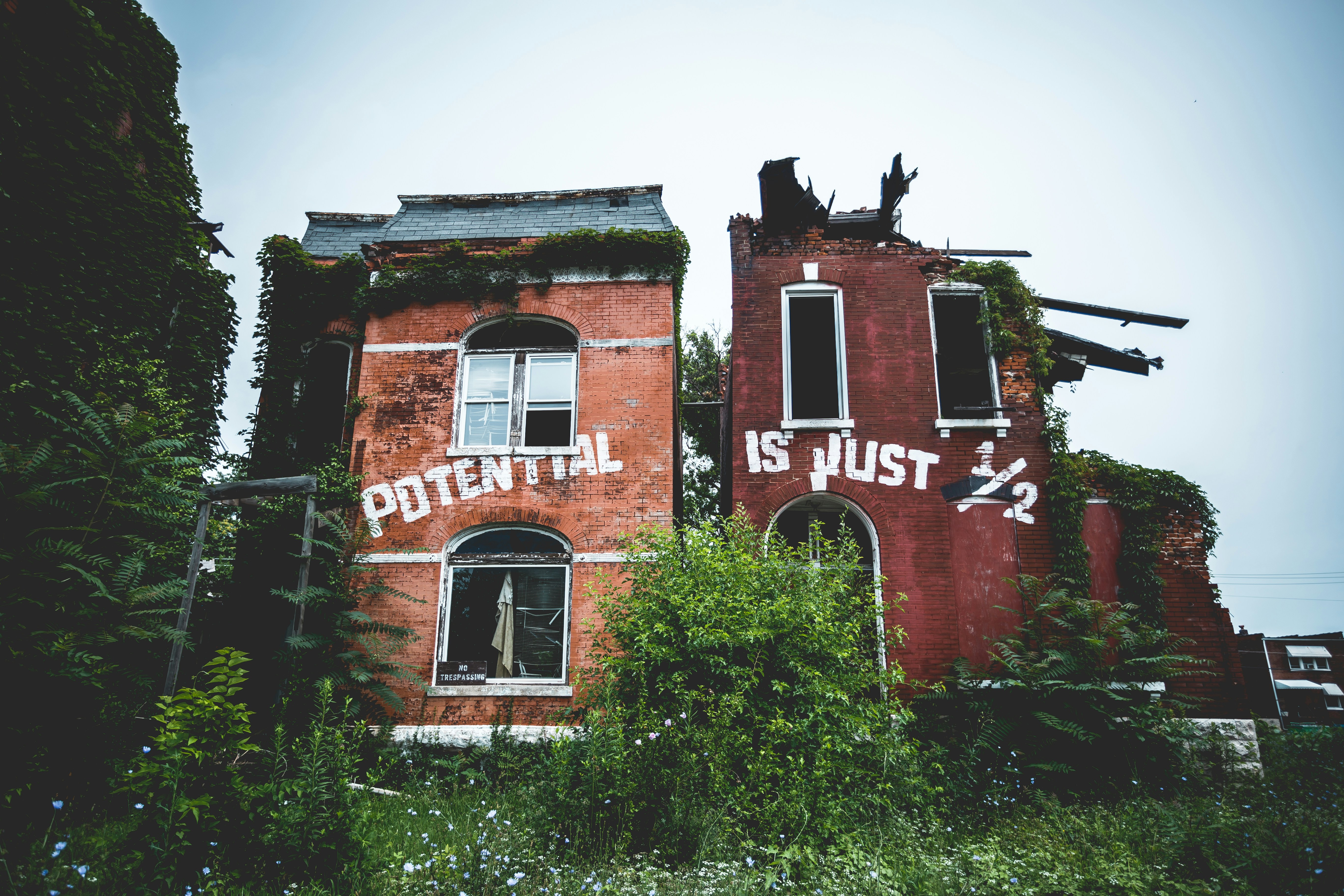 Abandoned brick building with graffiti reading 'Potential is just 1/2,' surrounded by overgrown greenery.