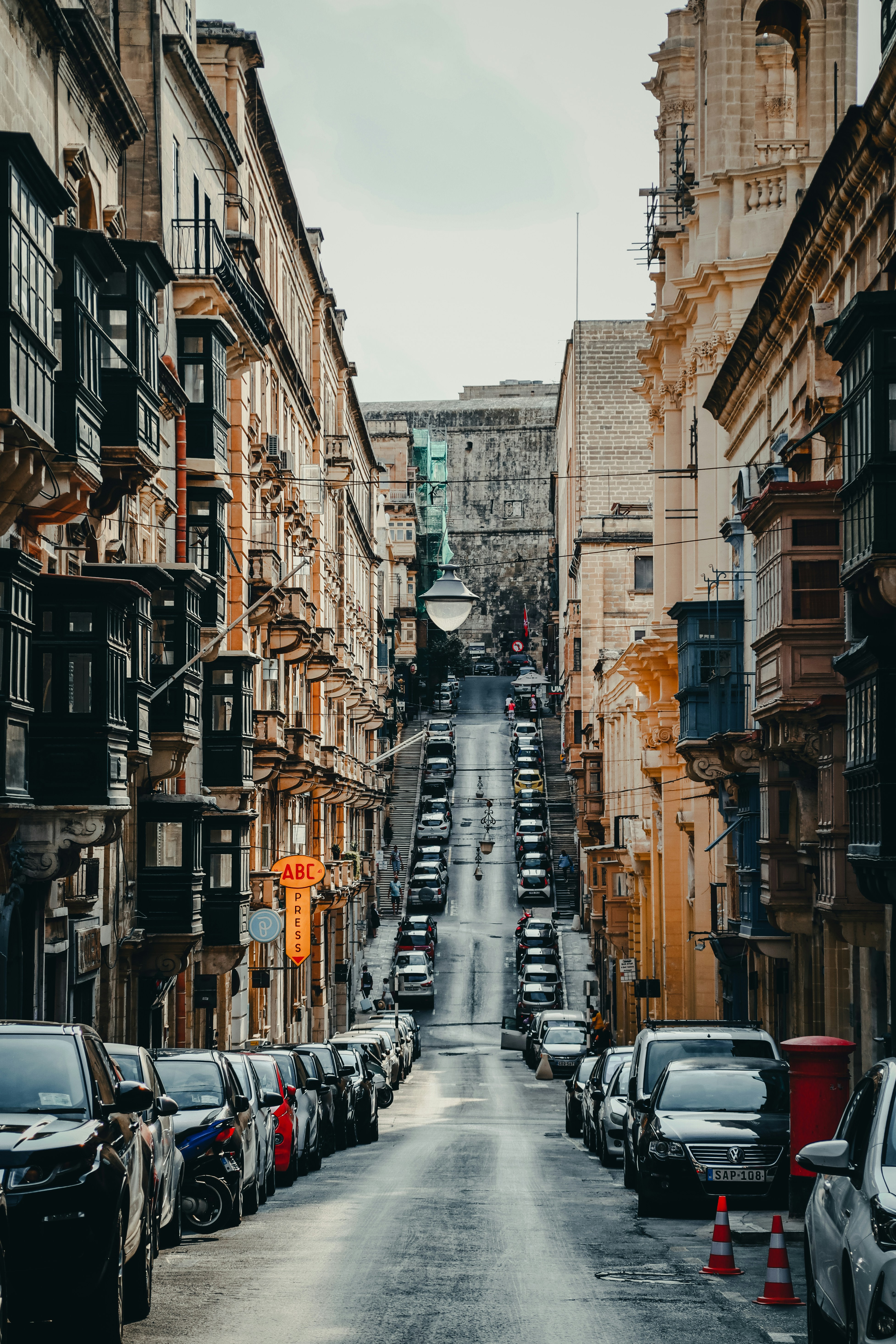 Narrow street lined with colorful balconies and parked cars, leading up a steep incline towards a historic fortification.